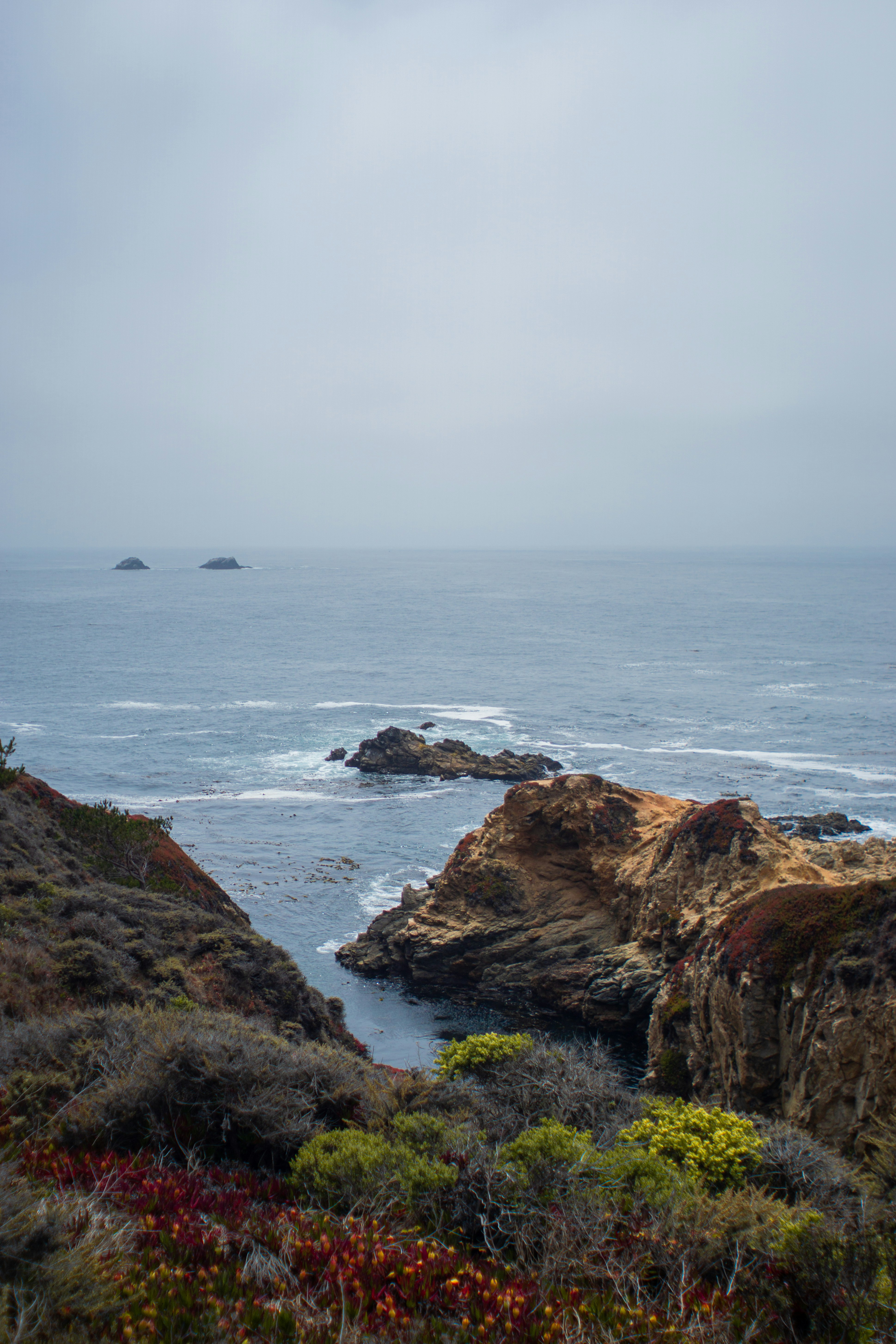 Rugged cliffs meet the tranquil ocean, with distant islands barely visible under a cloudy sky. Lush vegetation adds color to the coastal landscape.