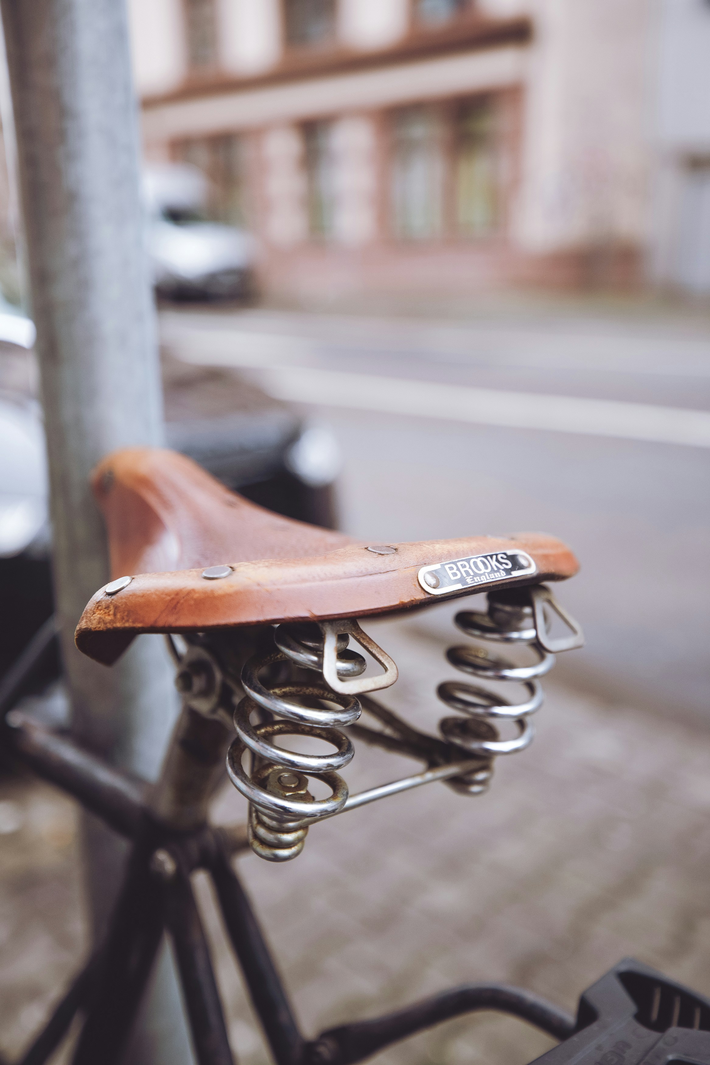 Close-up photograph of a leather Brooks bicycle saddle with visible springs, set against a blurred urban street backdrop.