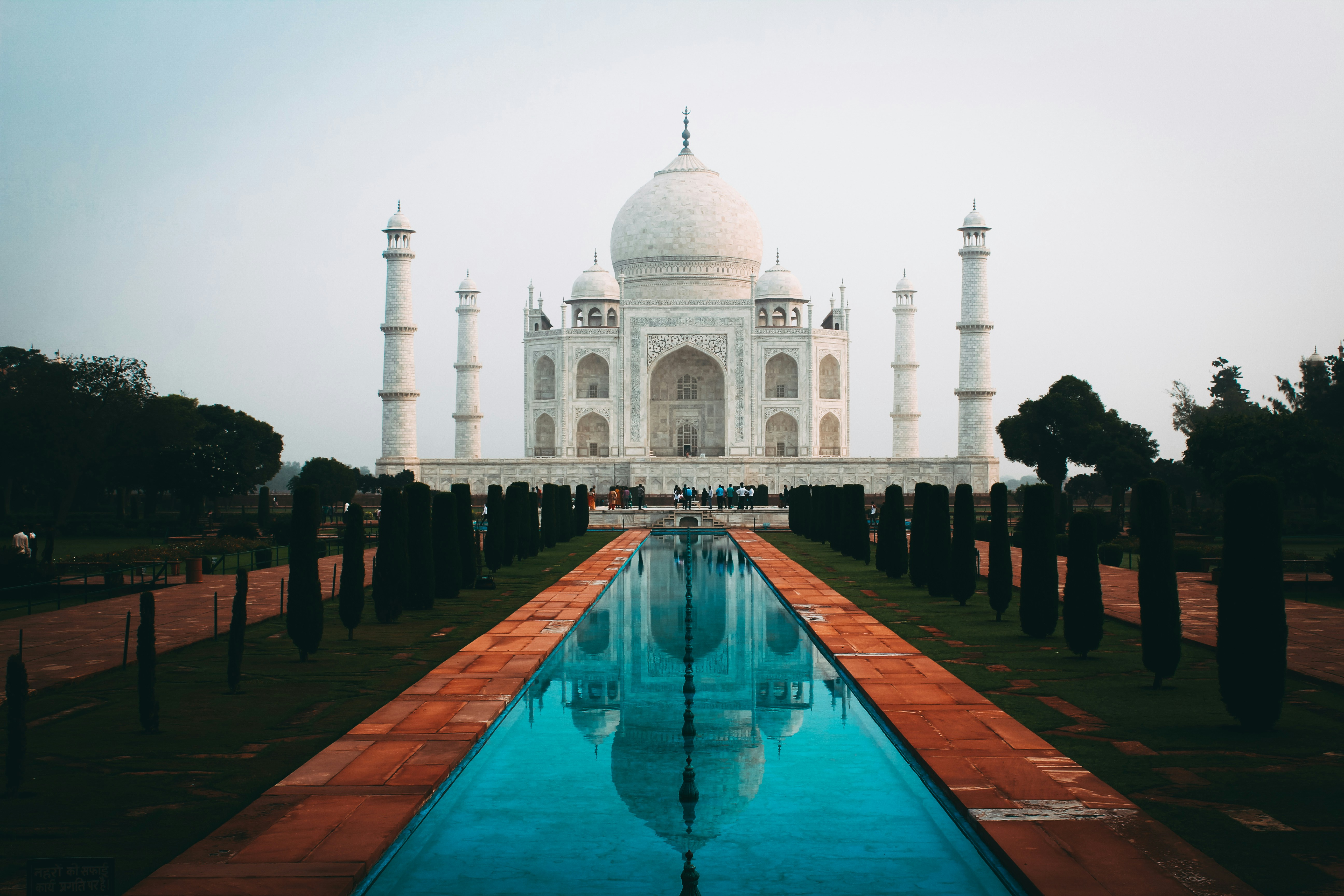 The Taj Mahal stands majestically, mirrored in a serene pool, surrounded by manicured gardens and towering trees.