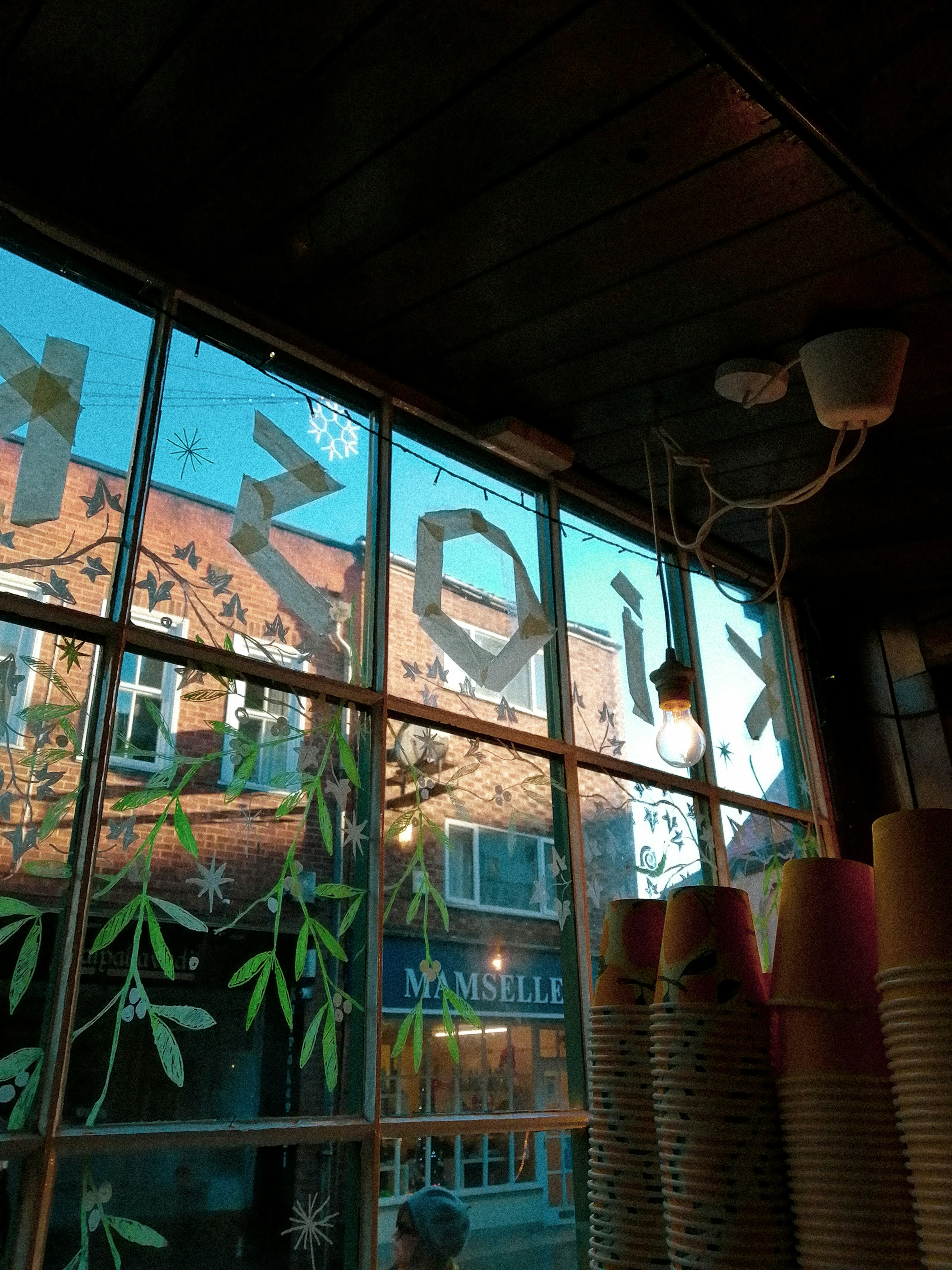 Sunlit café interior with large glass windows decorated with frosty shapes, stacked cups in the foreground, and a street scene visible outside.