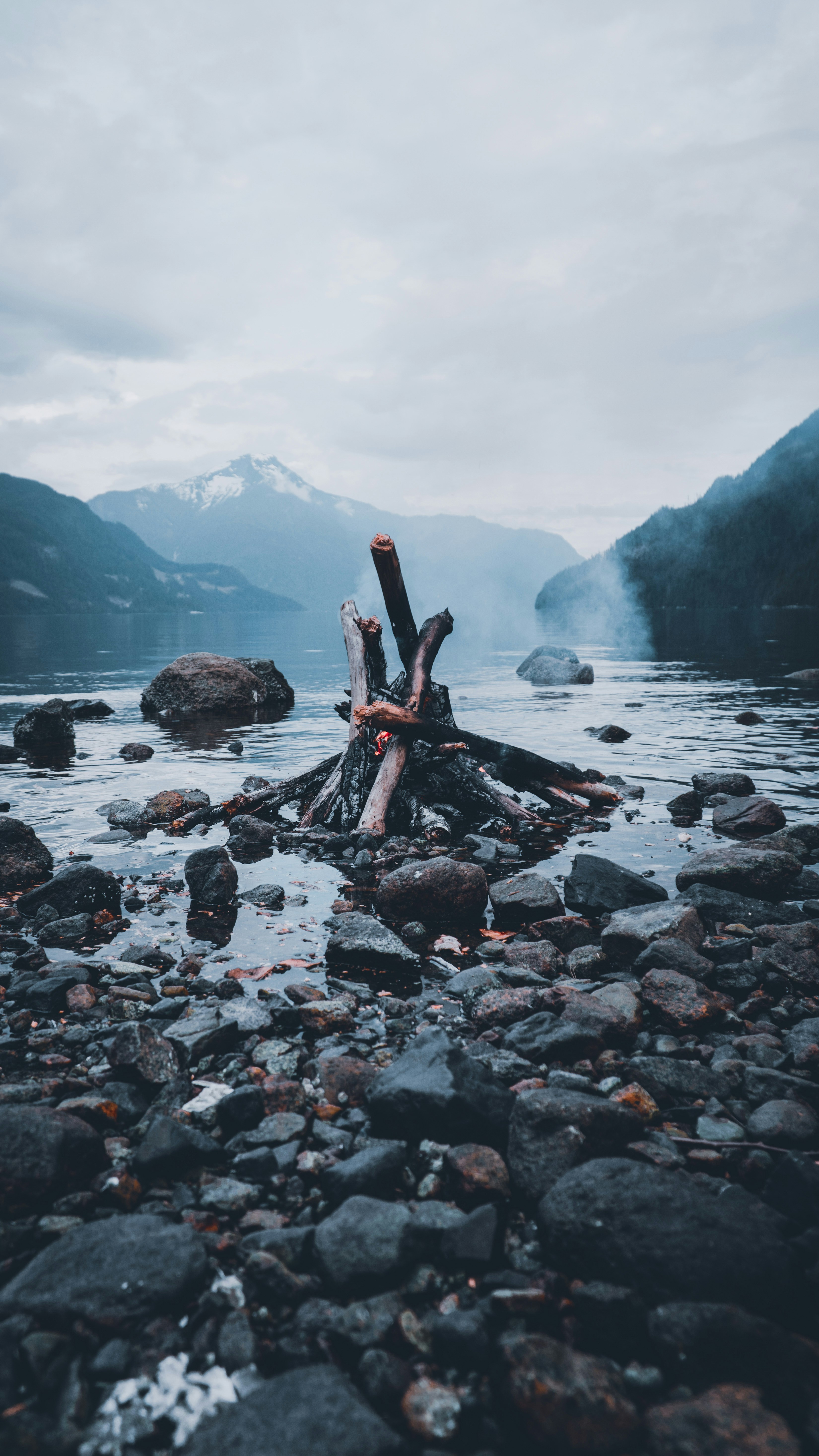 Campfire remnants surrounded by smooth stones and a tranquil lake, with mountains looming in the background. Smoke rises gently into the overcast sky.