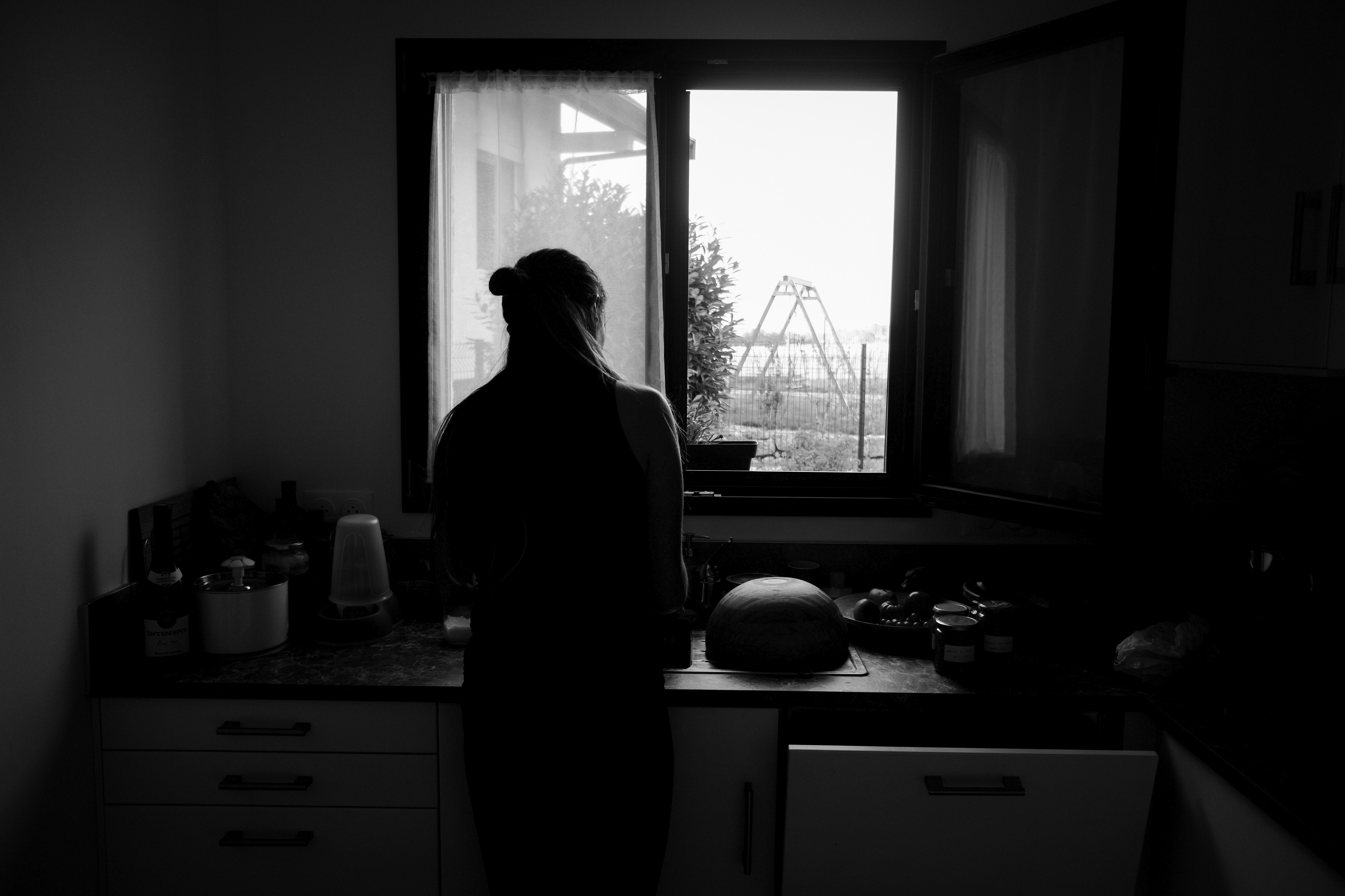 Silhouette of a person washing dishes in a dimly lit kitchen, with a view of a swing set outside the window. The scene evokes a sense of quiet solitude.