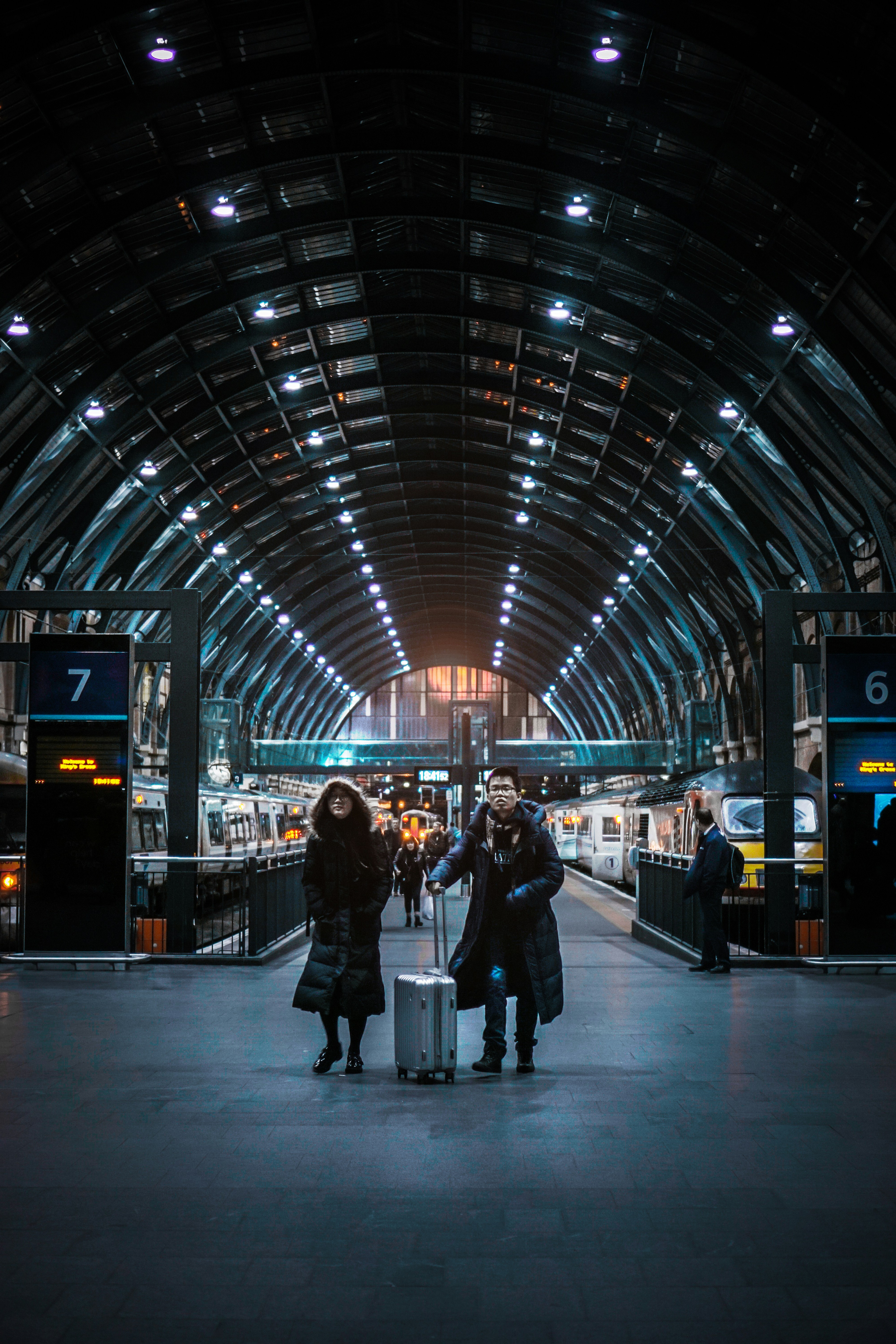 Two travelers stand beside a suitcase in a bustling train station, framed by a striking arched roof and illuminated by soft overhead lights.