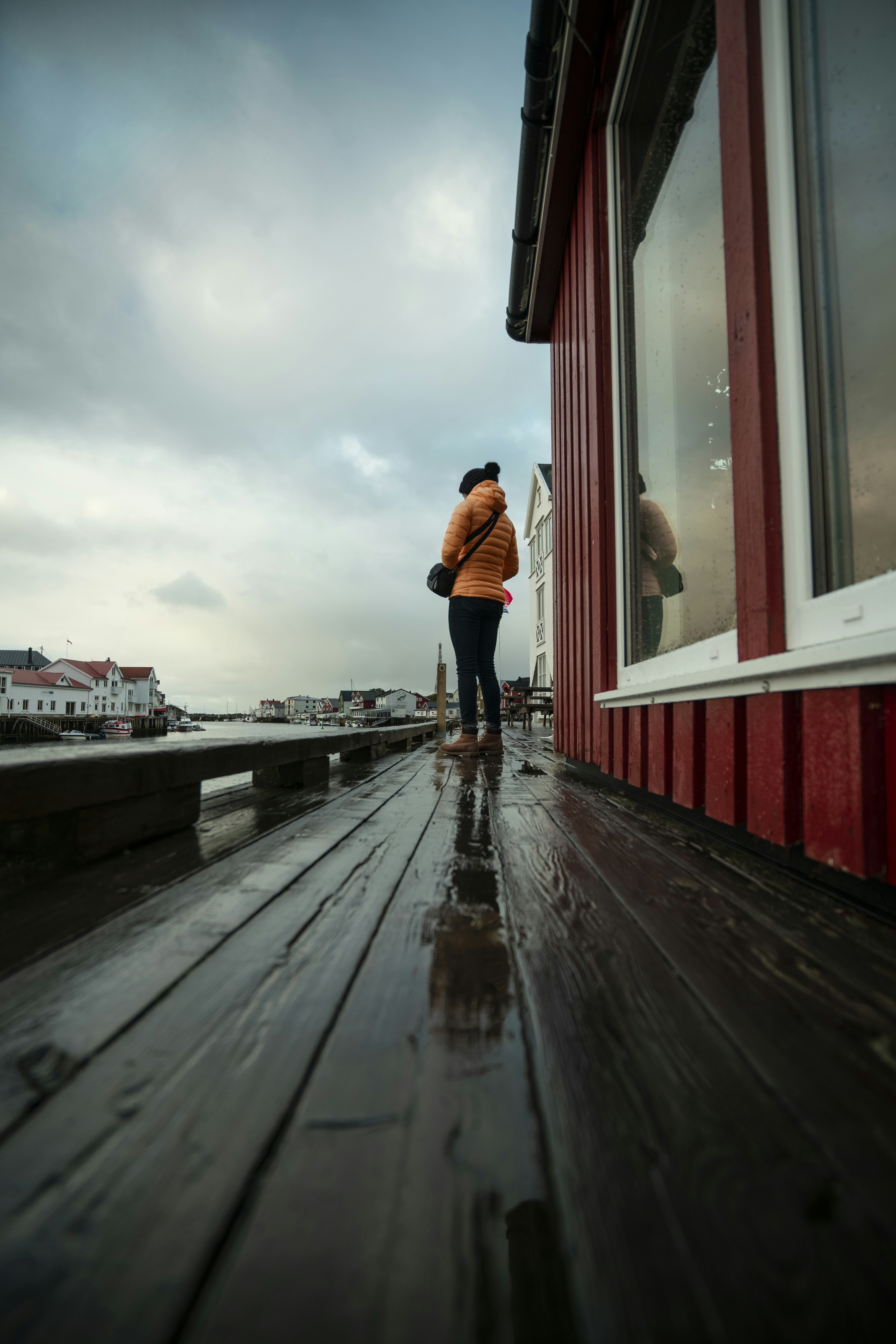 A person in an orange jacket stands pensively on a wet wooden walkway, reflecting the surrounding quaint architecture and cloudy skies.