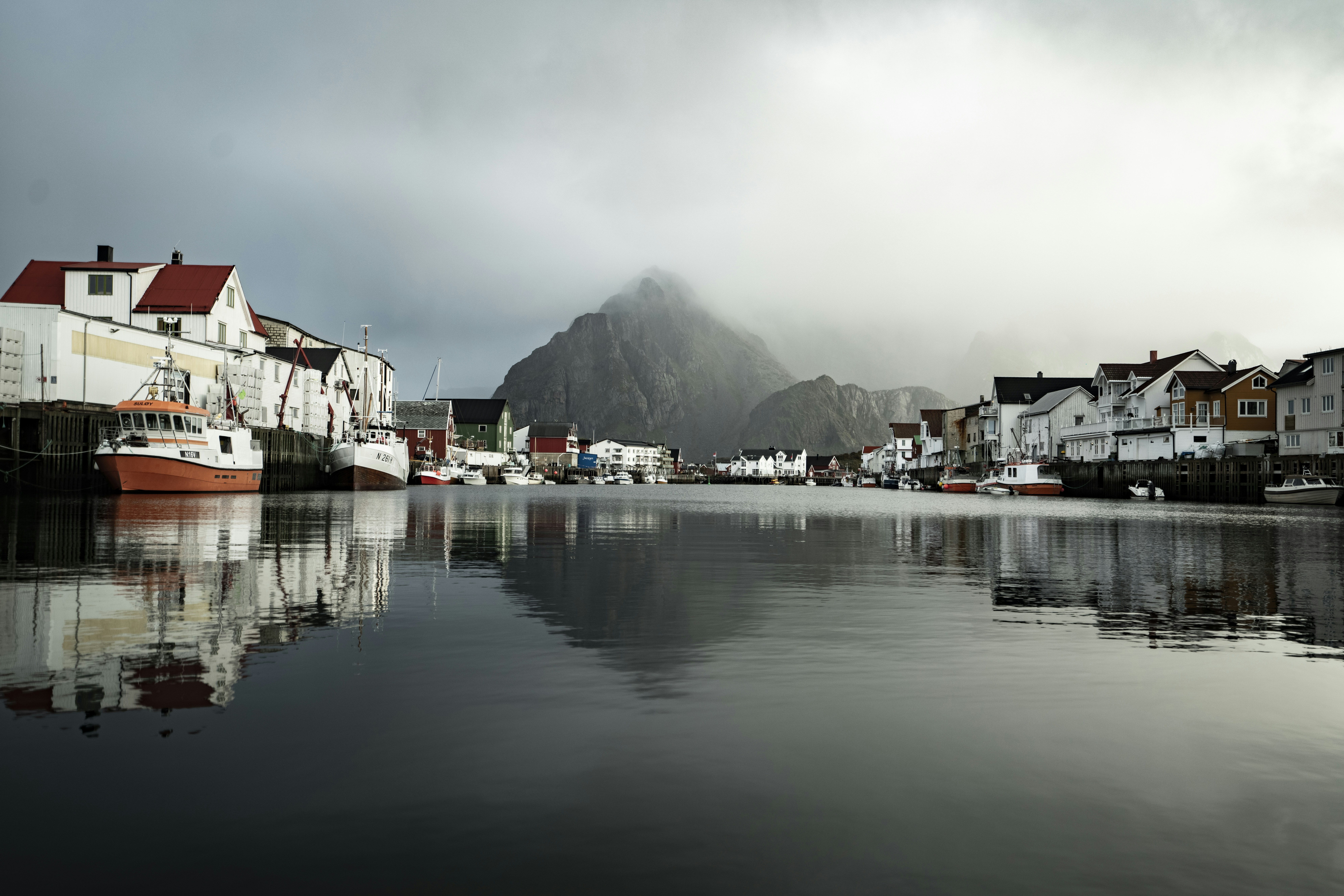 Fishing boats and quaint houses line a serene harbor, with misty mountains looming in the background. The water reflects the peaceful scene, creating a tranquil atmosphere.