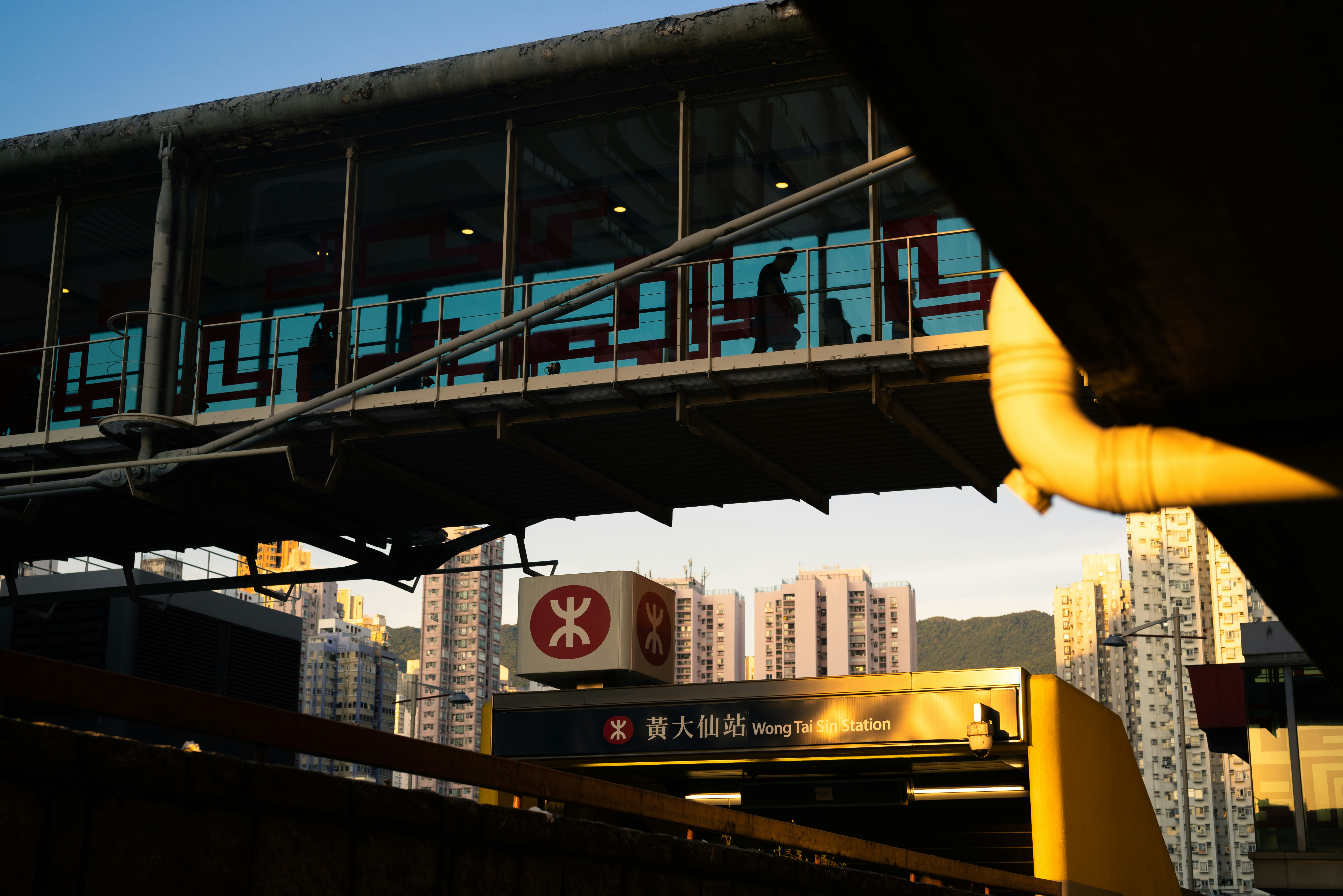 Silhouetted figures in a glass walkway with a city skyline backdrop at sunset.