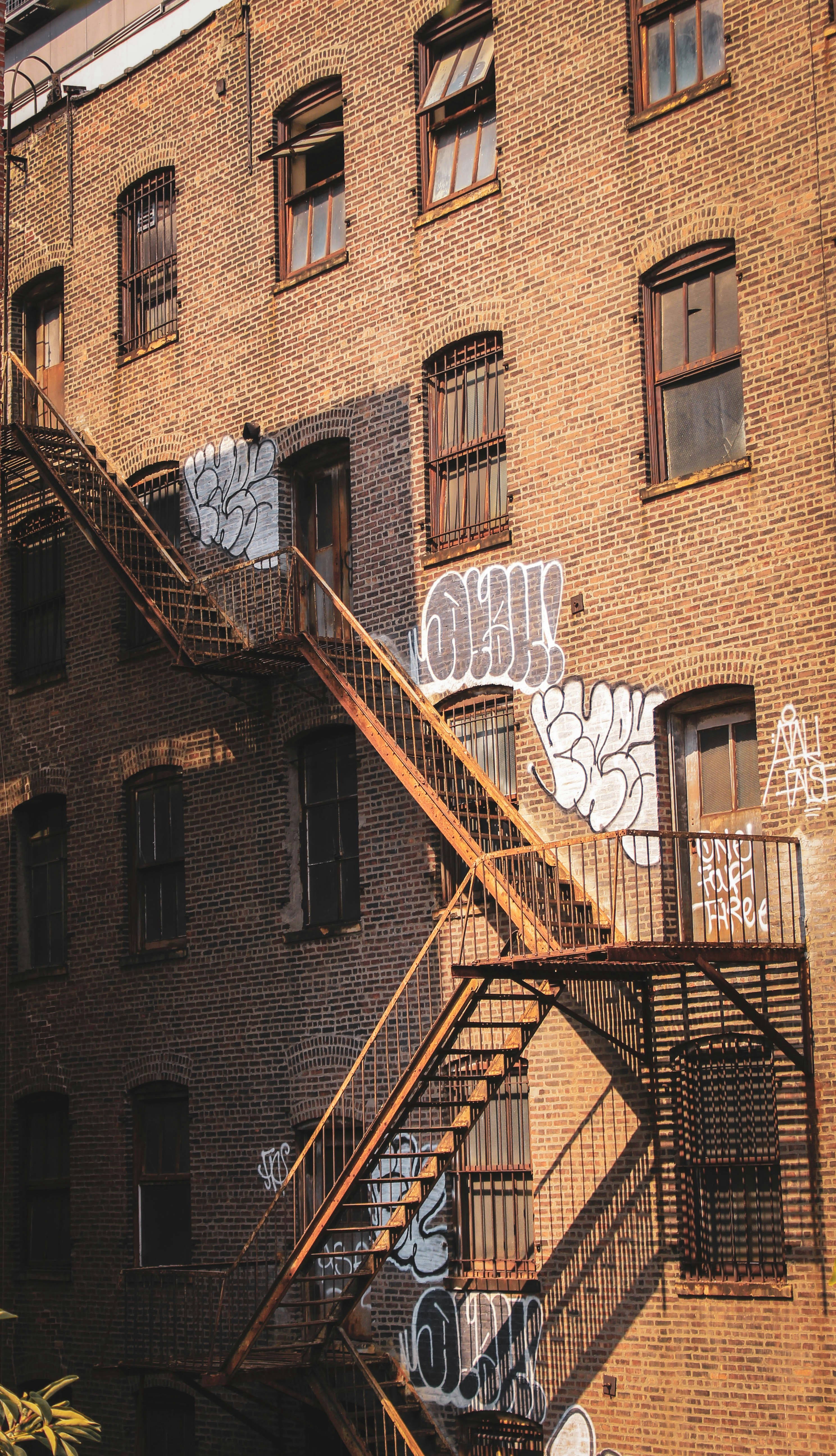 A weathered brick wall adorned with vibrant graffiti, featuring a prominent fire escape and a series of barred windows. The urban landscape reflects a blend of artistry and decay.