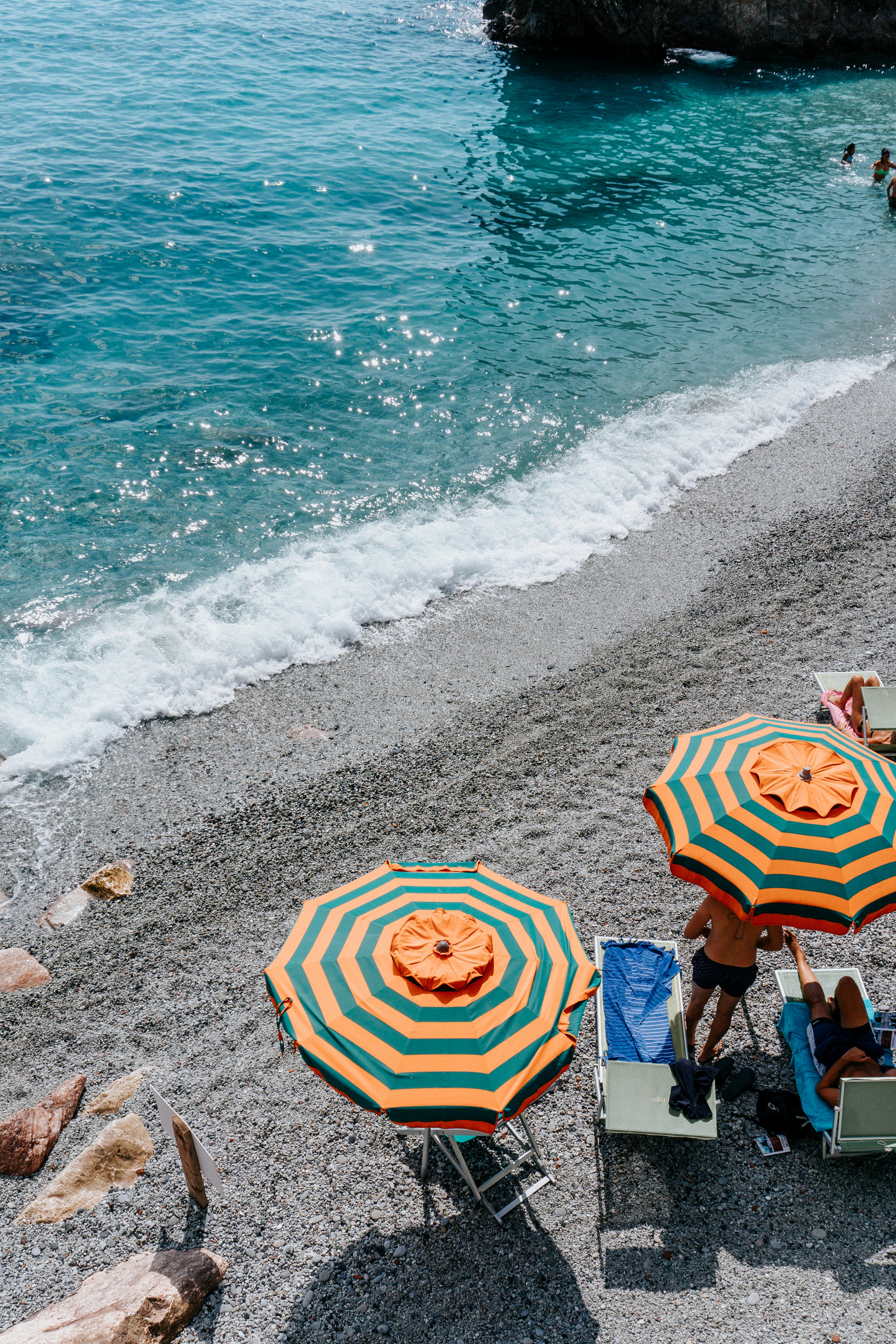 Colorful striped beach umbrellas provide shade for sunbathers on a pebbled beach beside clear turquoise waters.