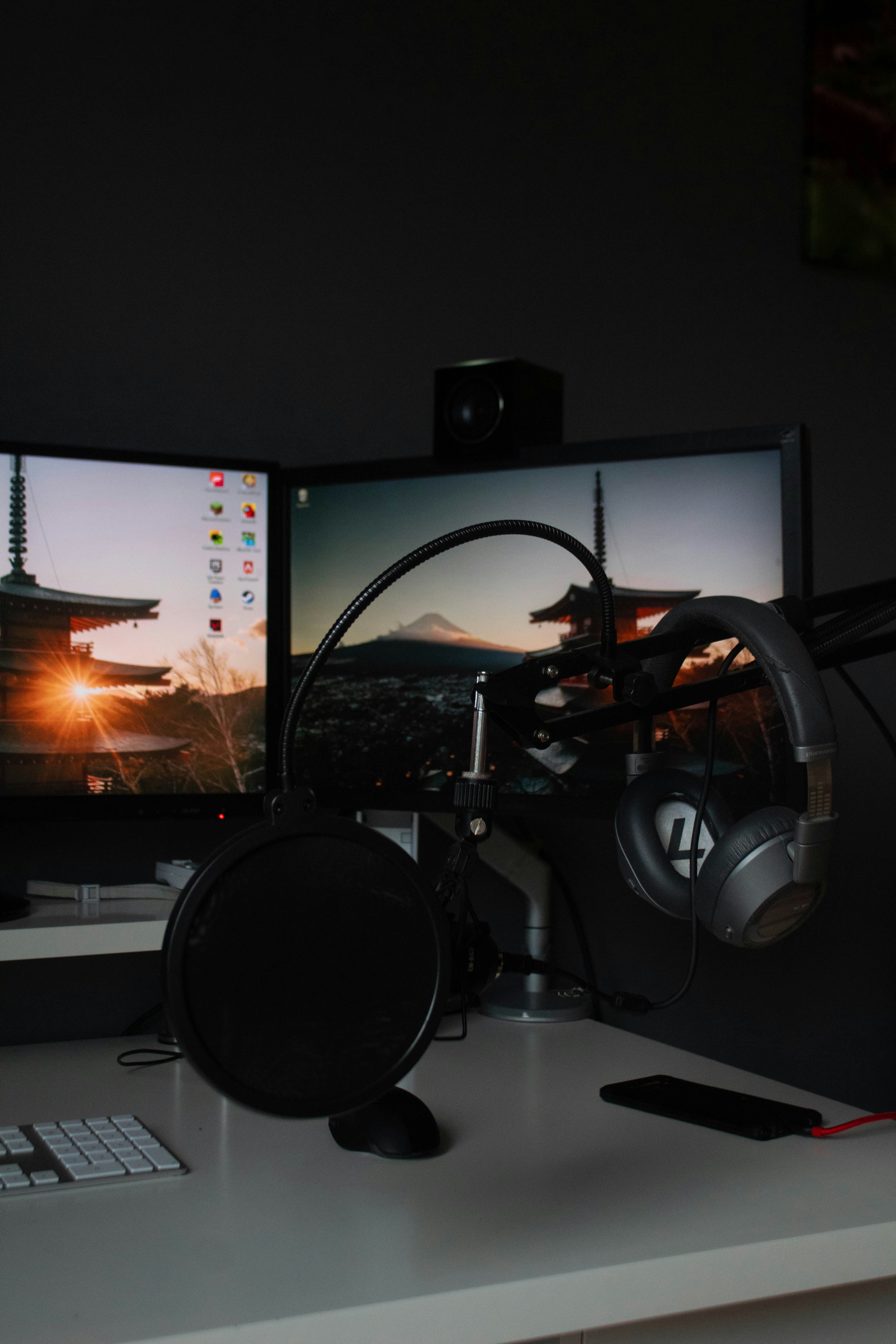 Dual-monitor workstation with a microphone, headphones, and keyboard on a clean desk setup. A phone rests beside the workspace, with a dark room backdrop emphasizing the illuminated screens.