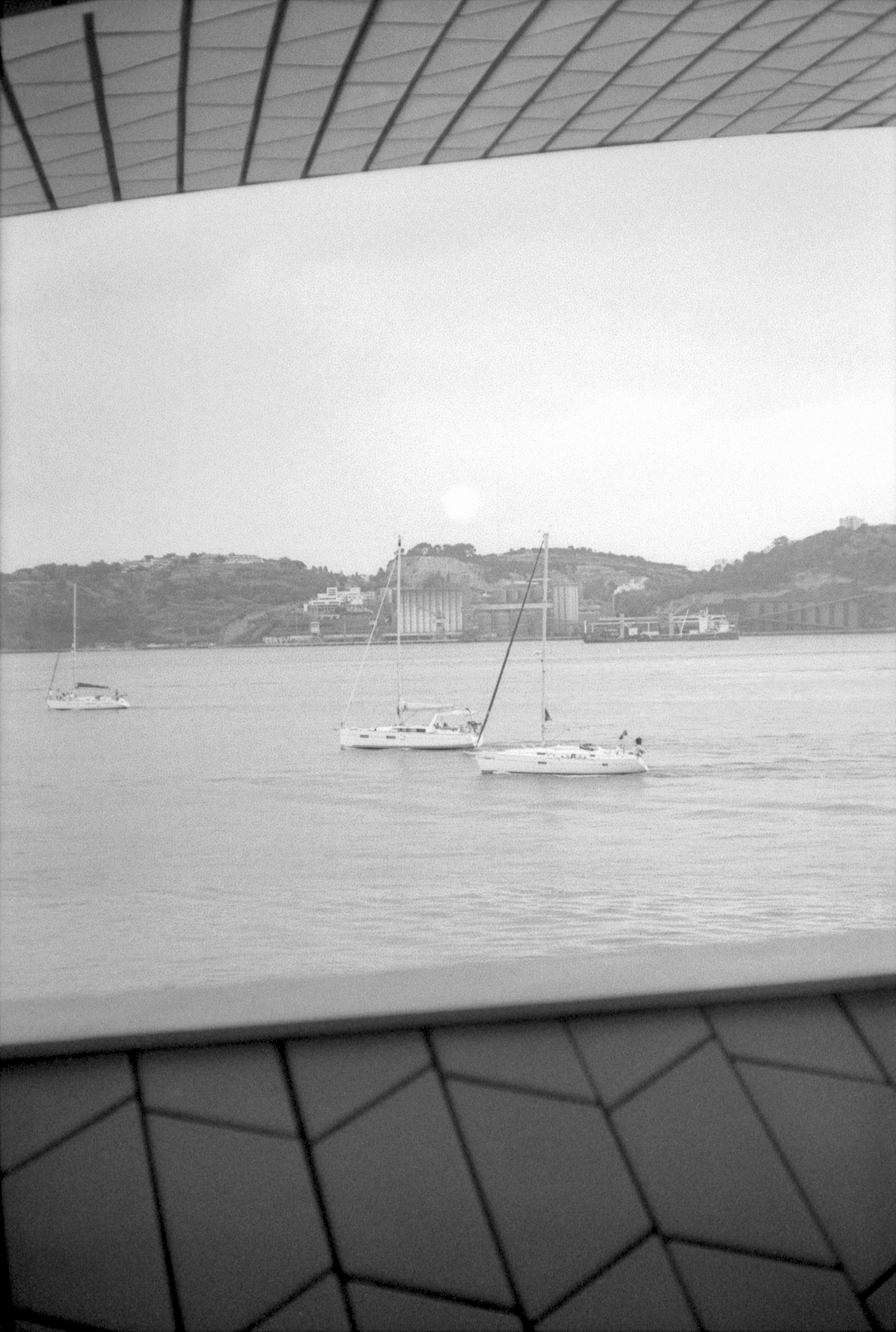 Black-and-white photograph of sailboats on a calm bay, viewed through angular foreground panels. Distant hills and shoreline create a muted backdrop.