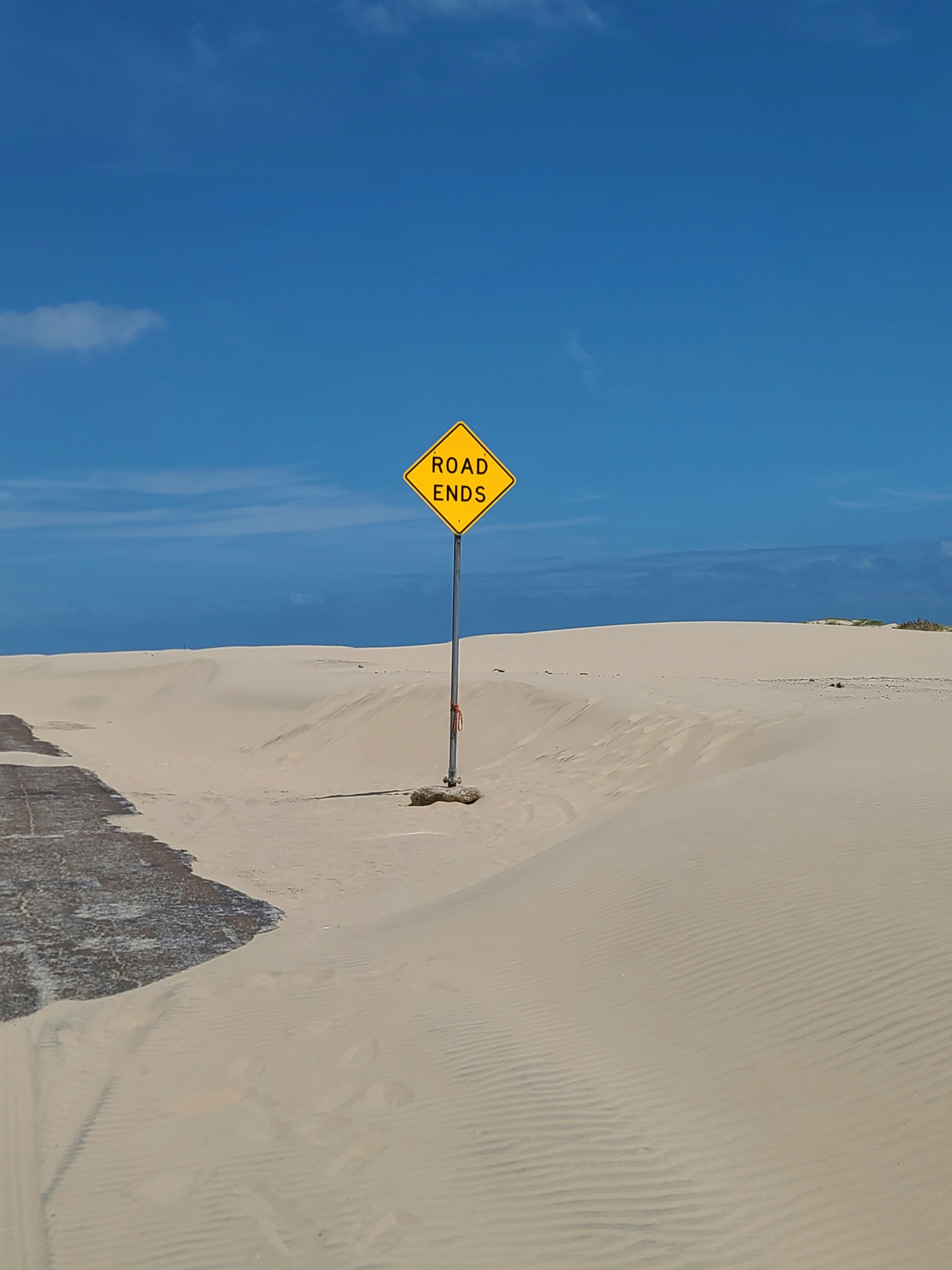 A yellow road-ends sign on a slender post rises from pale dunes beneath a vivid blue sky.