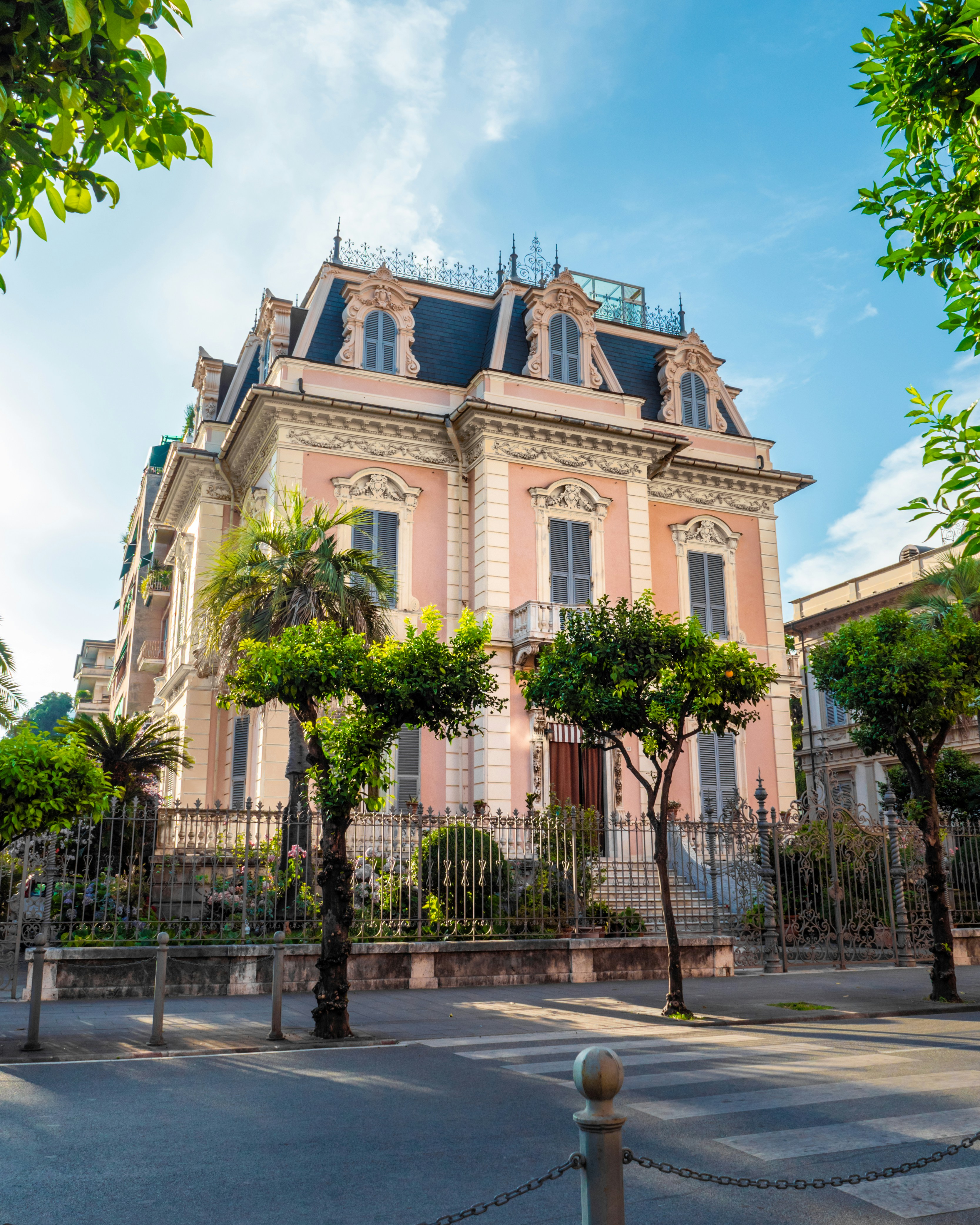 Ornate pink mansion surrounded by lush greenery and manicured gardens, showcasing exquisite architectural details. A tranquil urban scene unfolds under a clear blue sky.