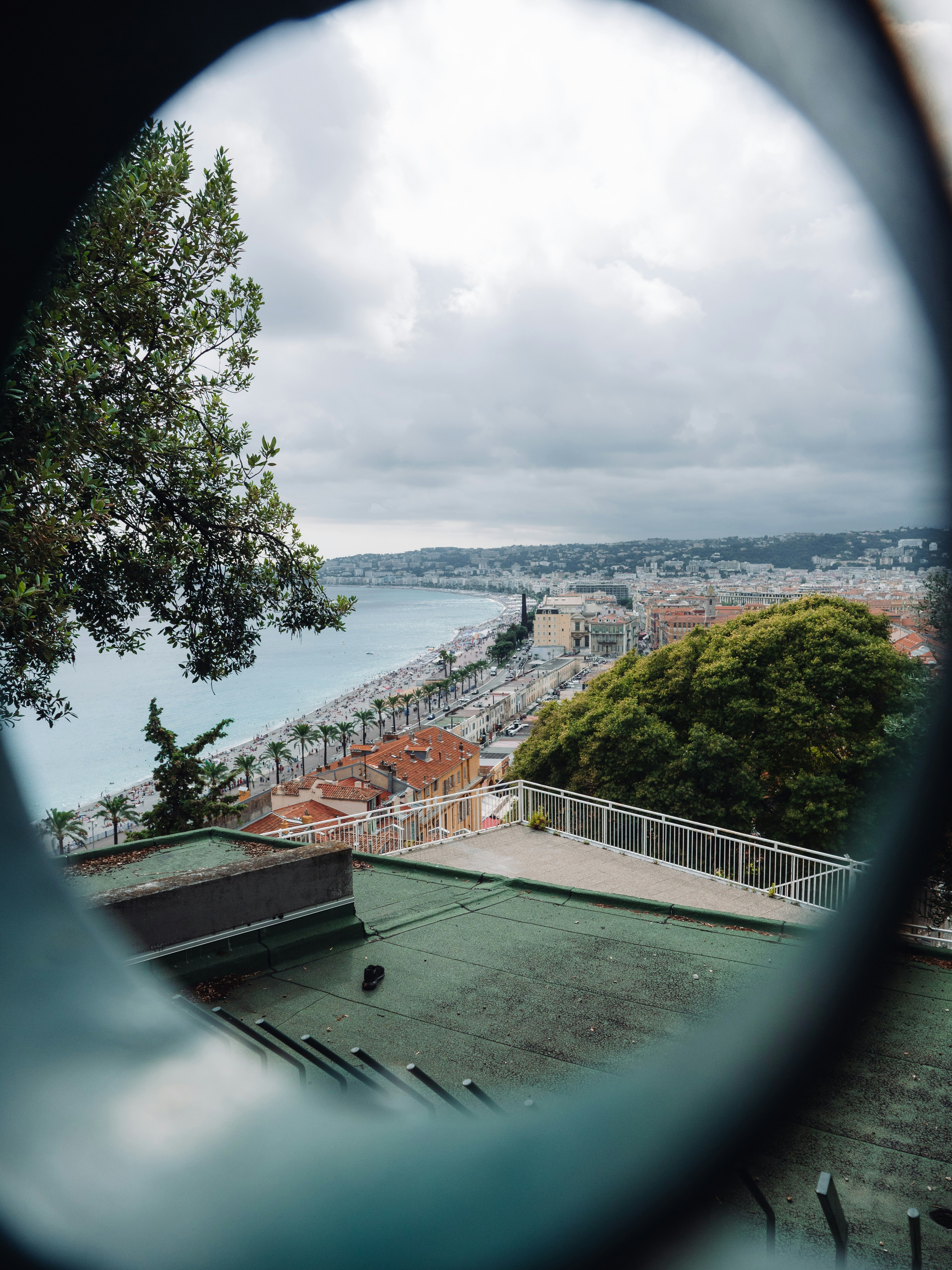 View of a coastal city framed by foliage, showcasing the shoreline and urban landscape under a cloudy sky.