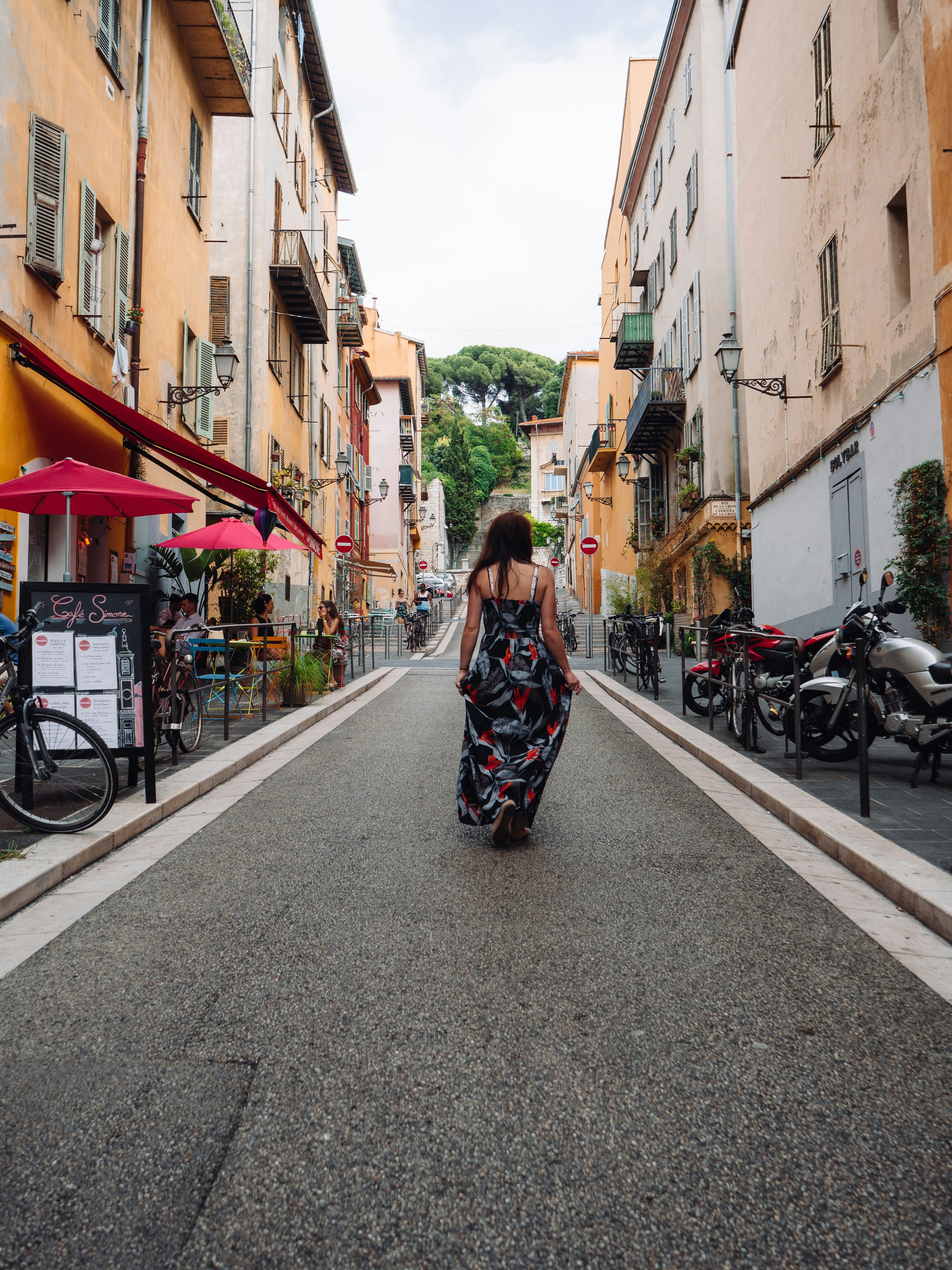 Woman in a floral dress walking down a narrow street lined with colorful buildings and bicycles. The atmosphere is lively with outdoor seating visible.