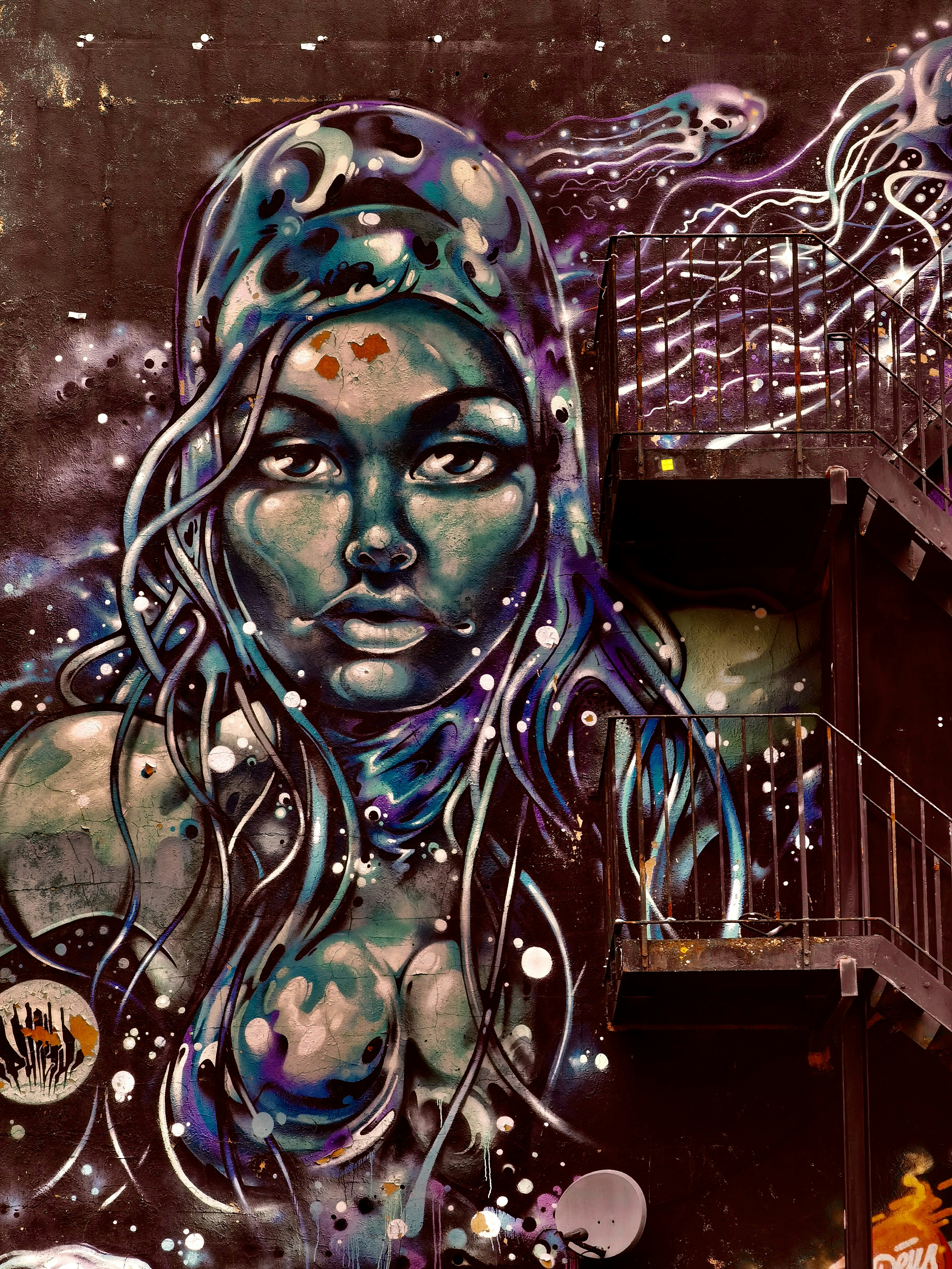Vibrant mural of a suspended-orbit woman painted on a brick wall, with color-rich nebula-like swirls and an external fire escape visible to the right. Artwork intended as street art or a large-scale illustration.