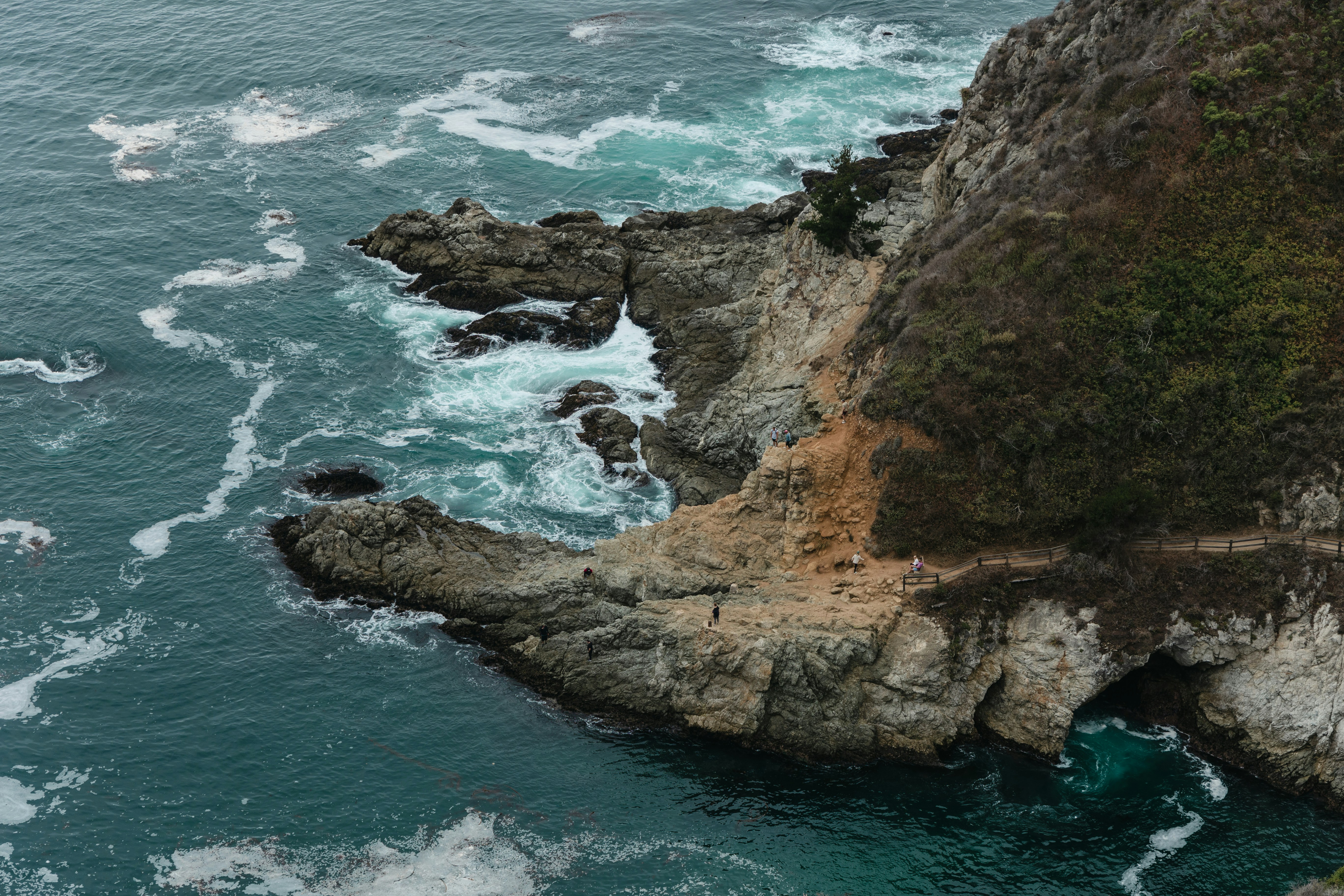 Rocky coastline meets turbulent waters, showcasing a blend of rugged terrain and vibrant ocean waves. A winding path invites exploration along the shore.