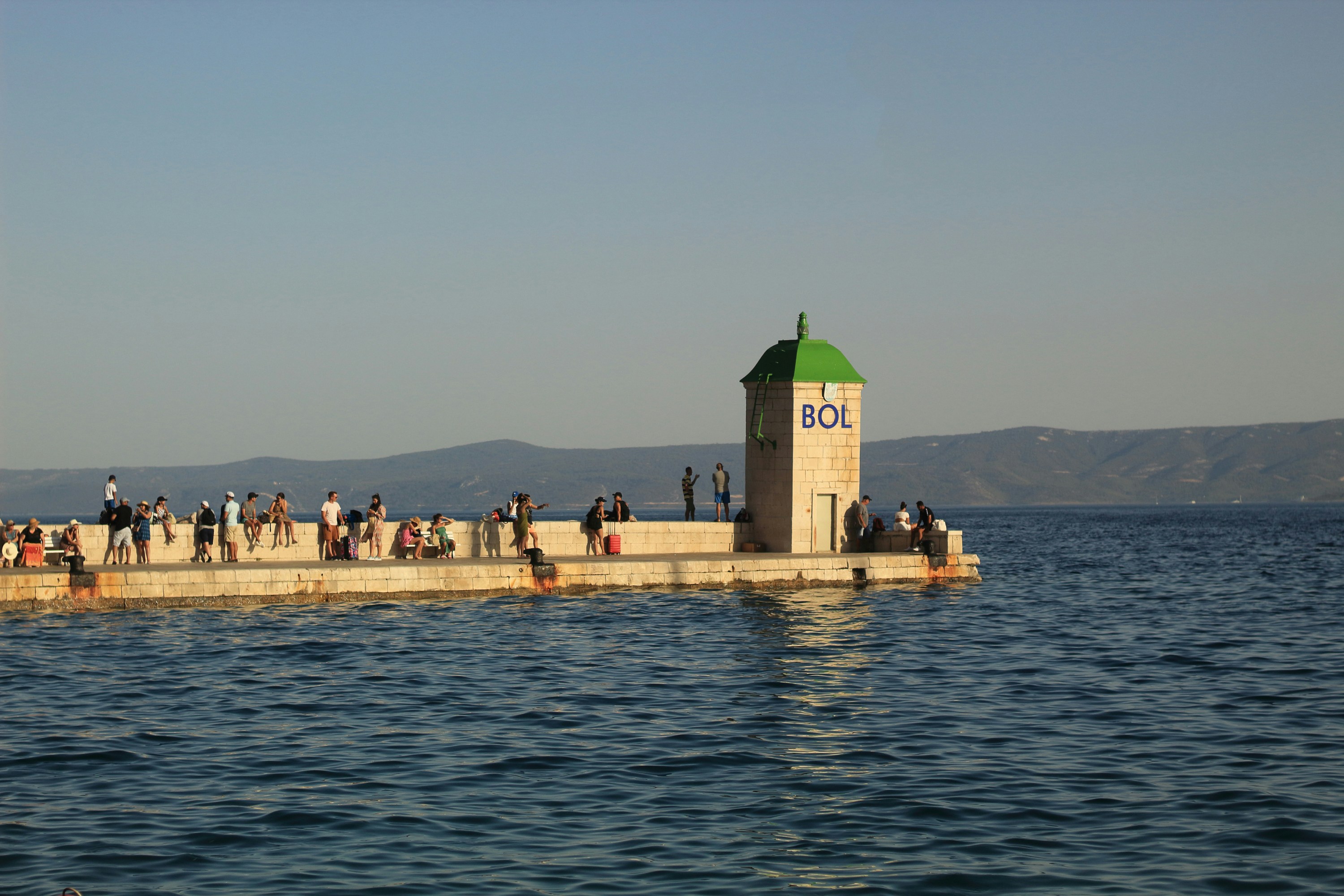 People gather on a pier with a small tower, surrounded by calm waters under a clear sky.