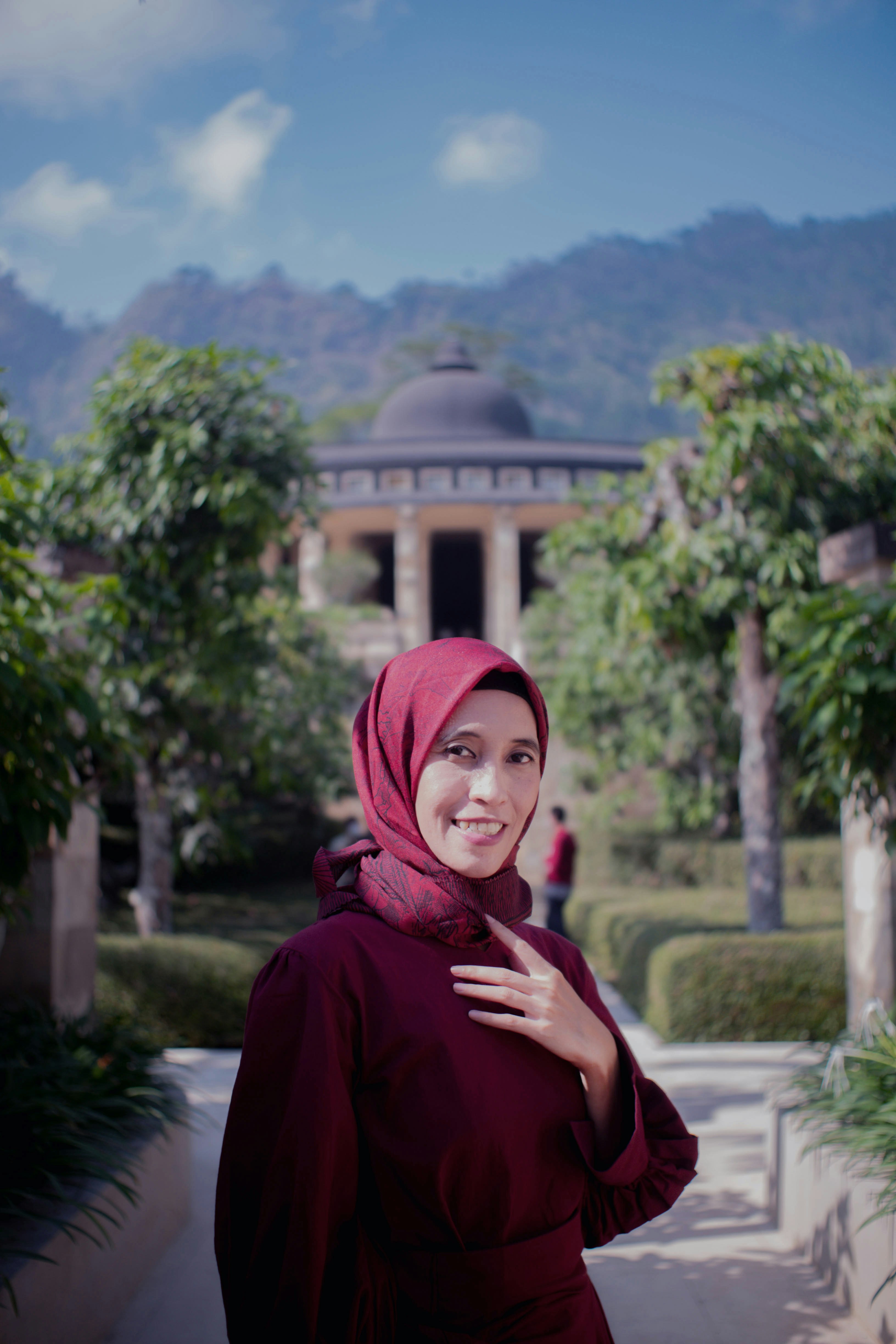 A woman in a maroon hijab stands gracefully in a lush garden, framed by verdant foliage and distant hills. The architectural structure in the background adds depth to the serene atmosphere.