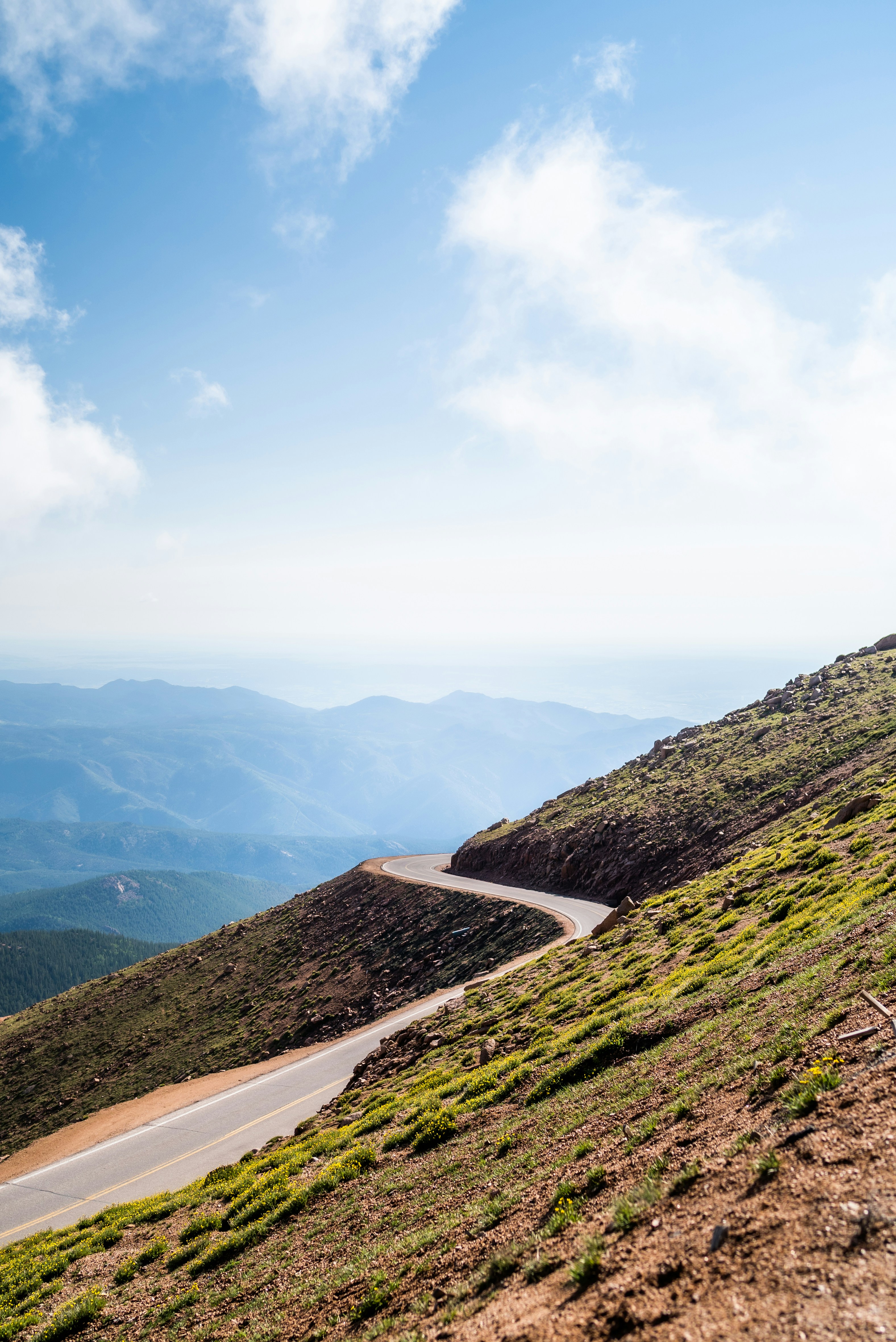 A winding road curves along a mountainside, offering a glimpse of distant peaks under a clear blue sky. Lush greenery contrasts with the rugged terrain.