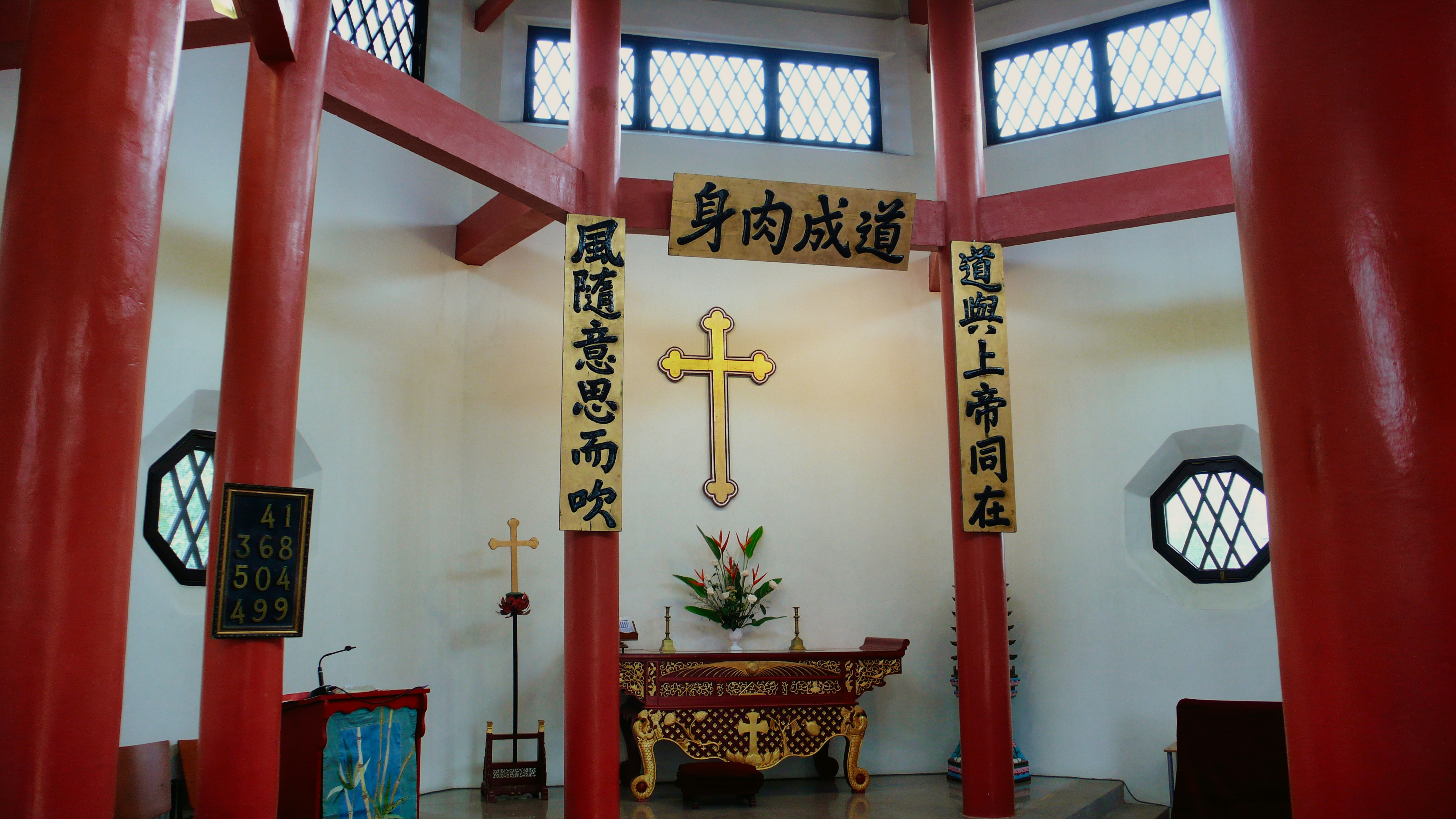 Interior photograph of a shrine-like space, with a golden cross above an ornate altar. Crimson columns and hanging banners frame the scene.