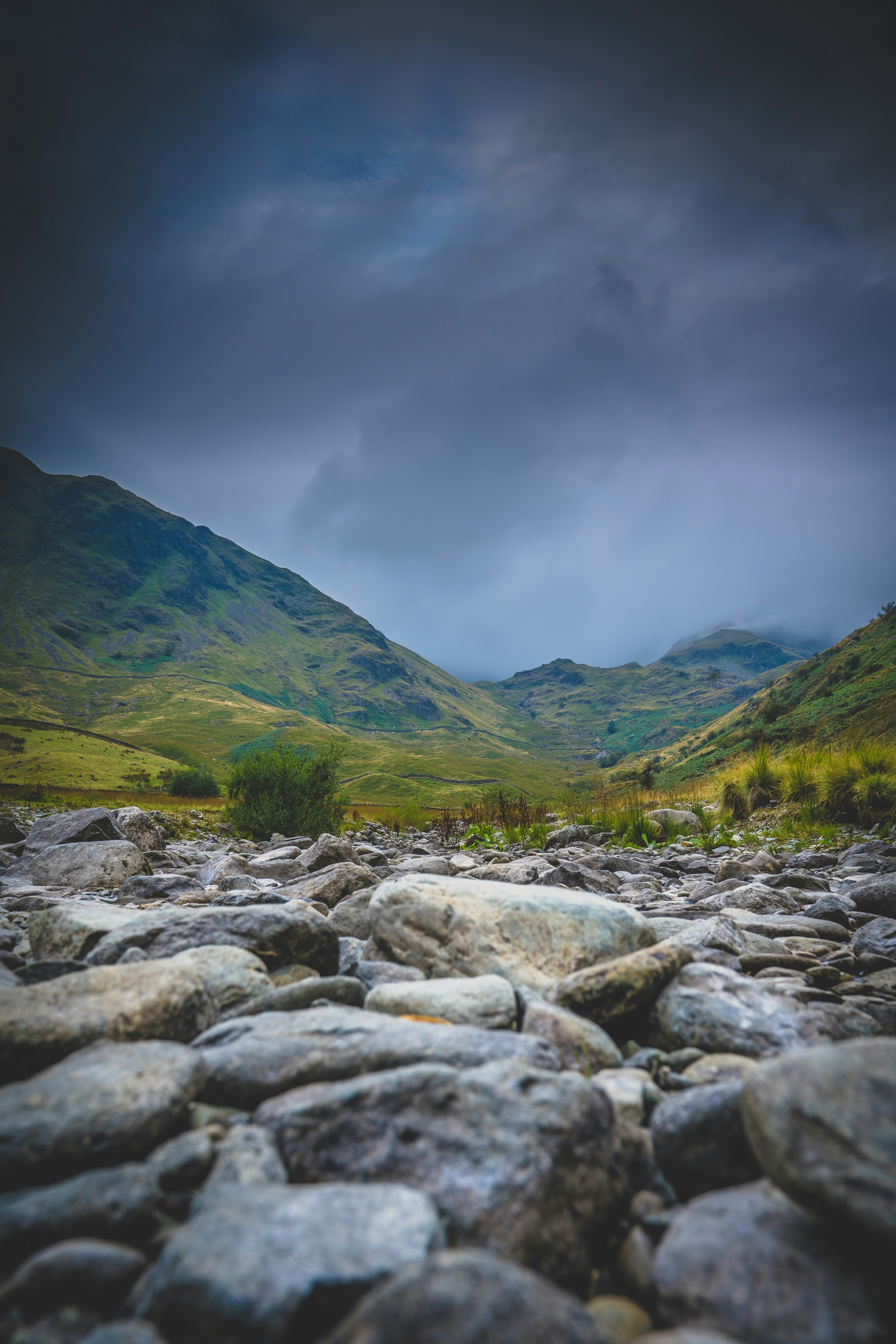 Rocky riverbed leading into a lush valley with towering mountains under a moody sky. The scene captures the rugged beauty of nature.