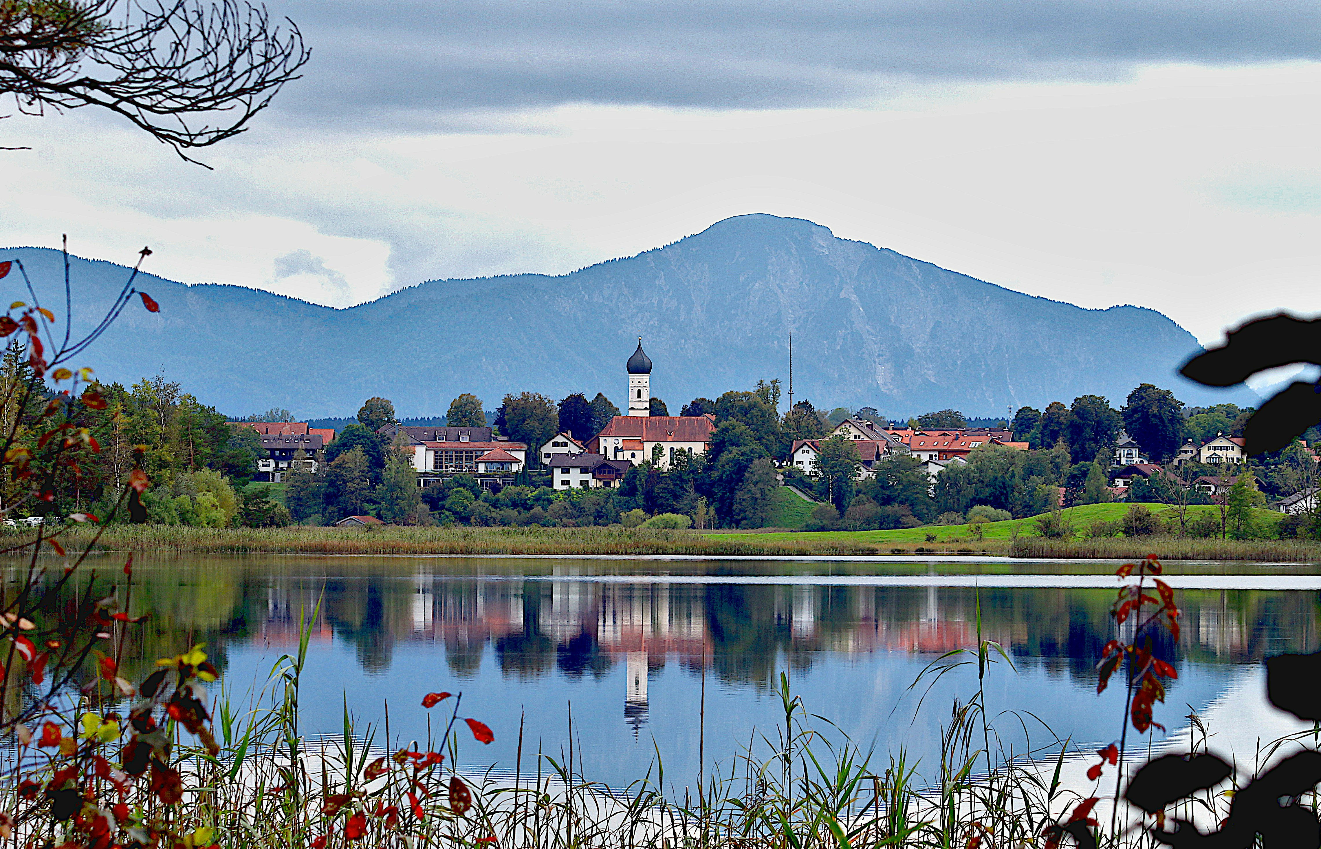 Lakeside village with a church steeple nestled against a backdrop of mountains, reflecting in calm waters. Vibrant foliage frames the scene.