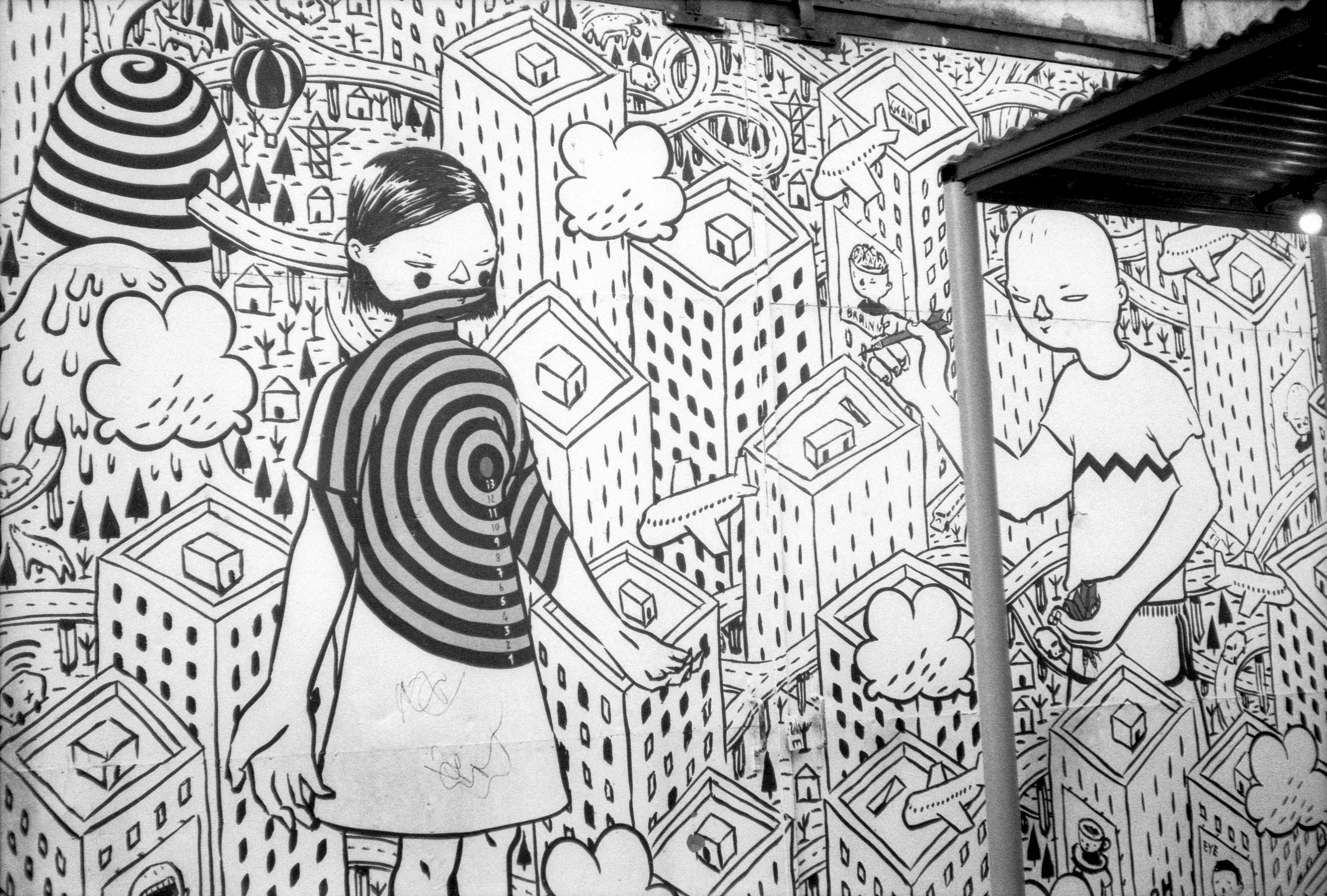 Monochrome mural illustration of a striped-clad figure navigating a city of tall, cubist buildings and whimsical clouds. The scene reads as a playful urban drawing.