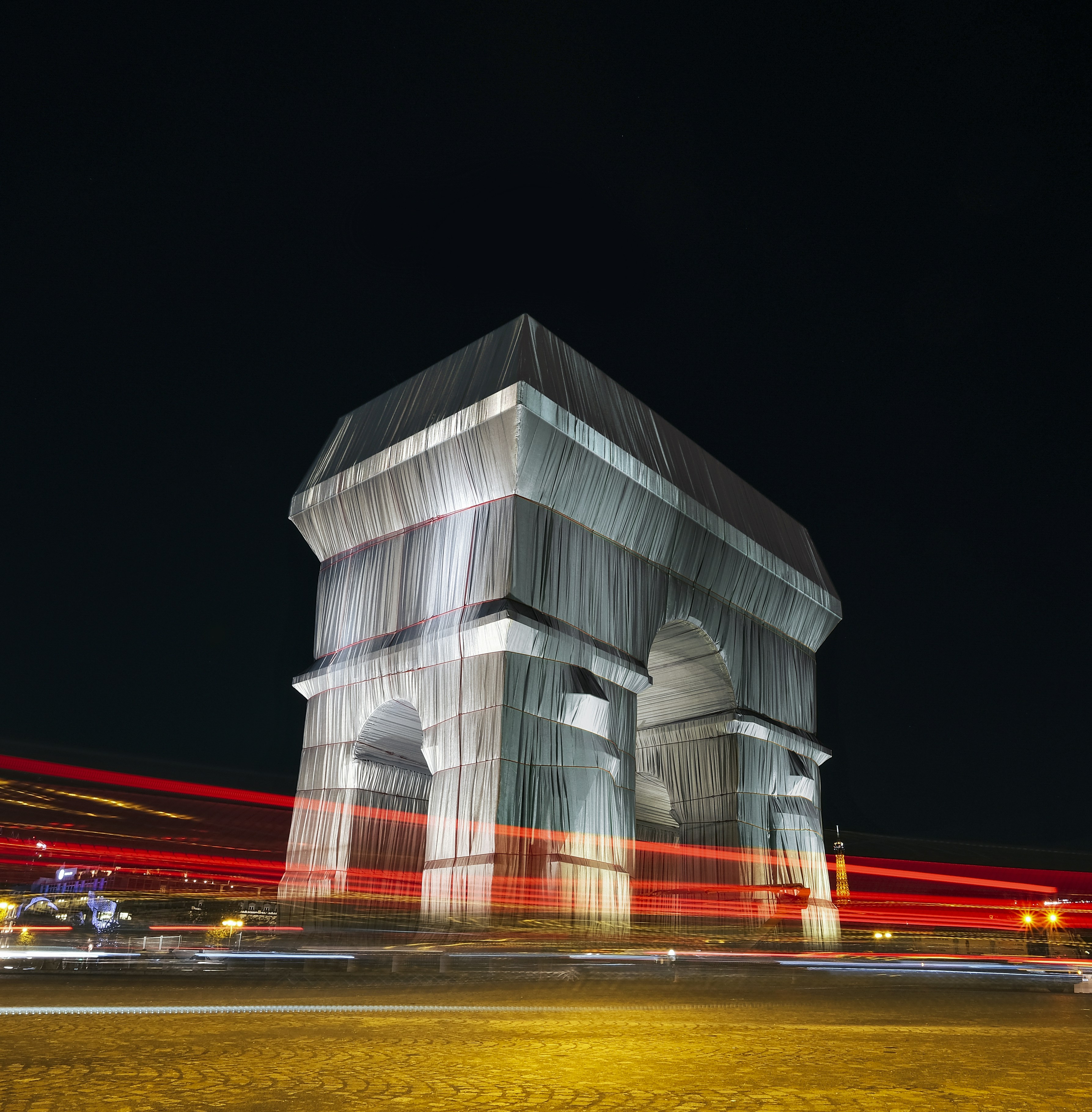 The Arc de Triomphe enveloped in fabric, illuminated against the night sky, with streaks of light from passing vehicles. 