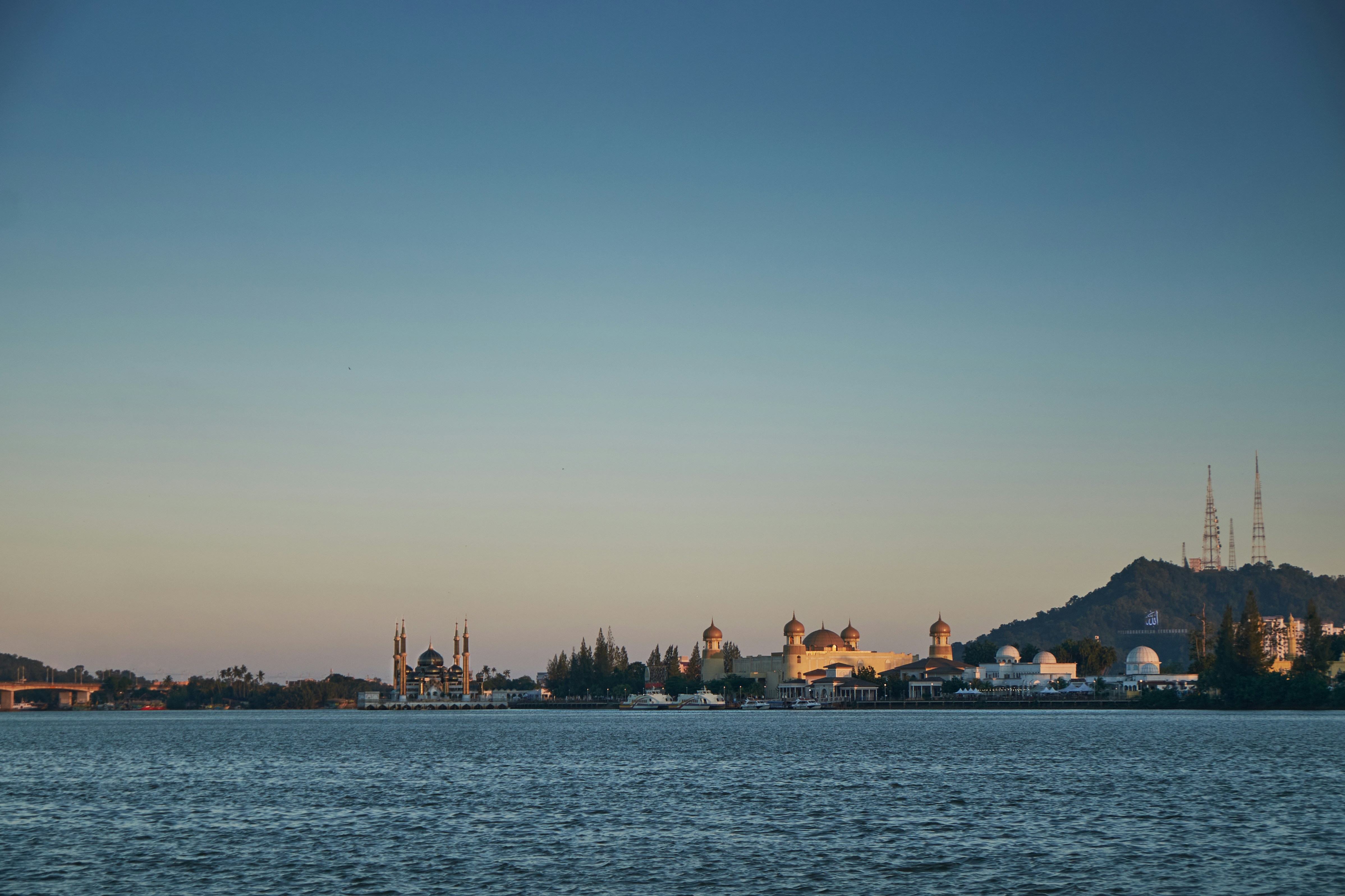 Calm waters with a city skyline and distant hills under a clear evening sky.