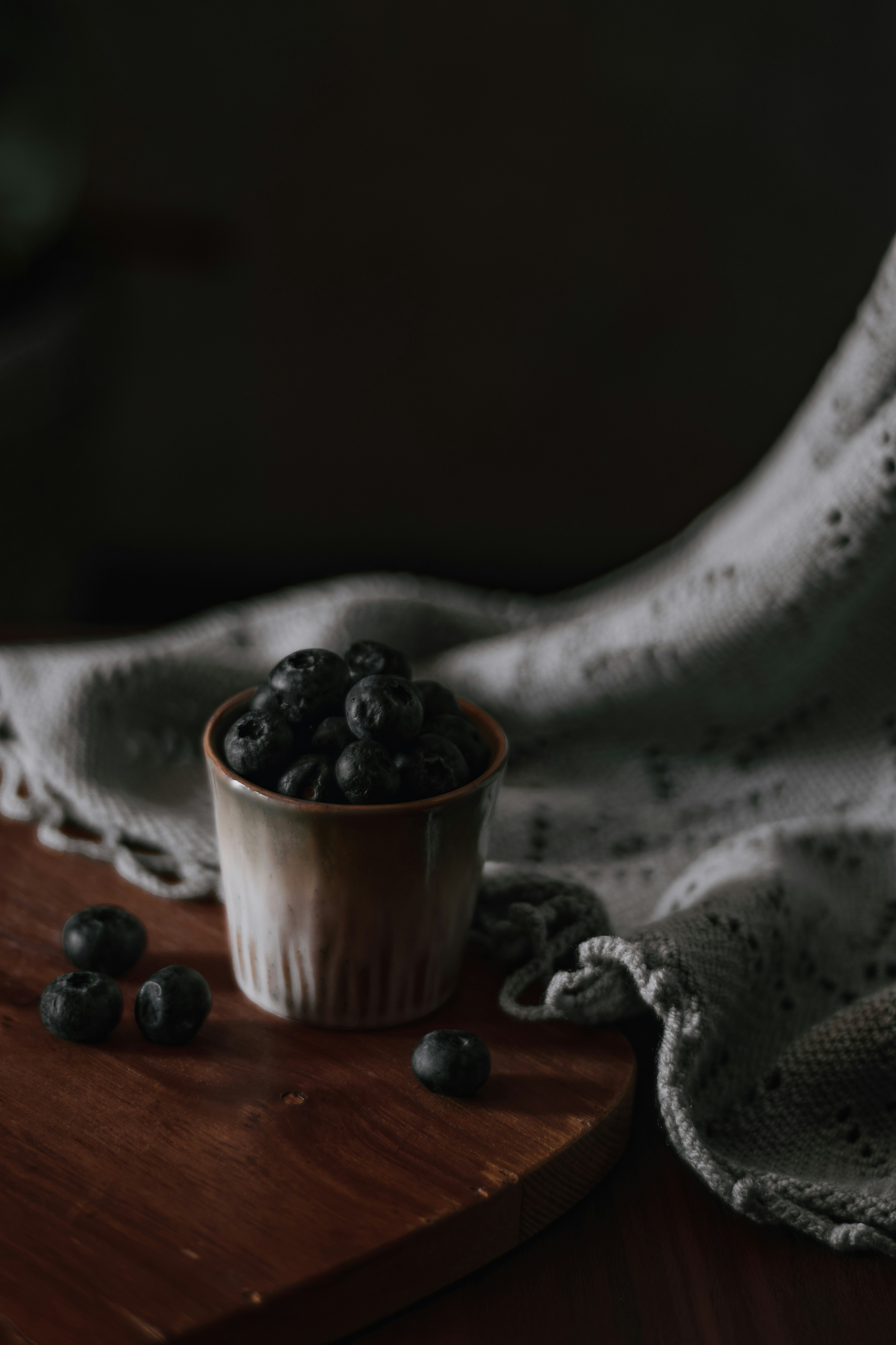 A rustic bowl filled with fresh blueberries rests on a wooden surface, accompanied by scattered berries and a soft, textured blanket in the background.