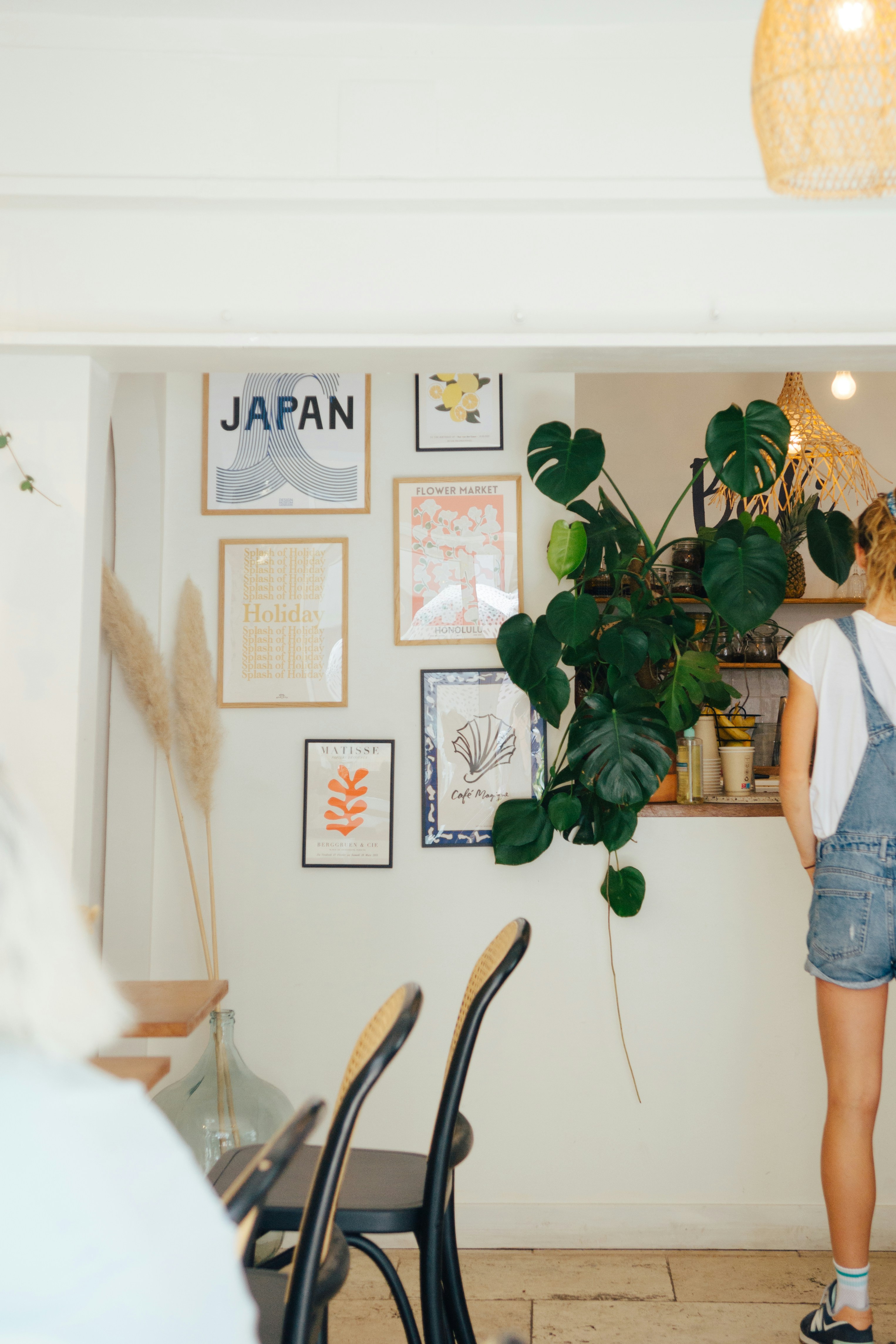 A serene café corner adorned with vibrant artwork and lush greenery, showcasing a relaxed atmosphere. A woman stands near the plants, engaging with the space.