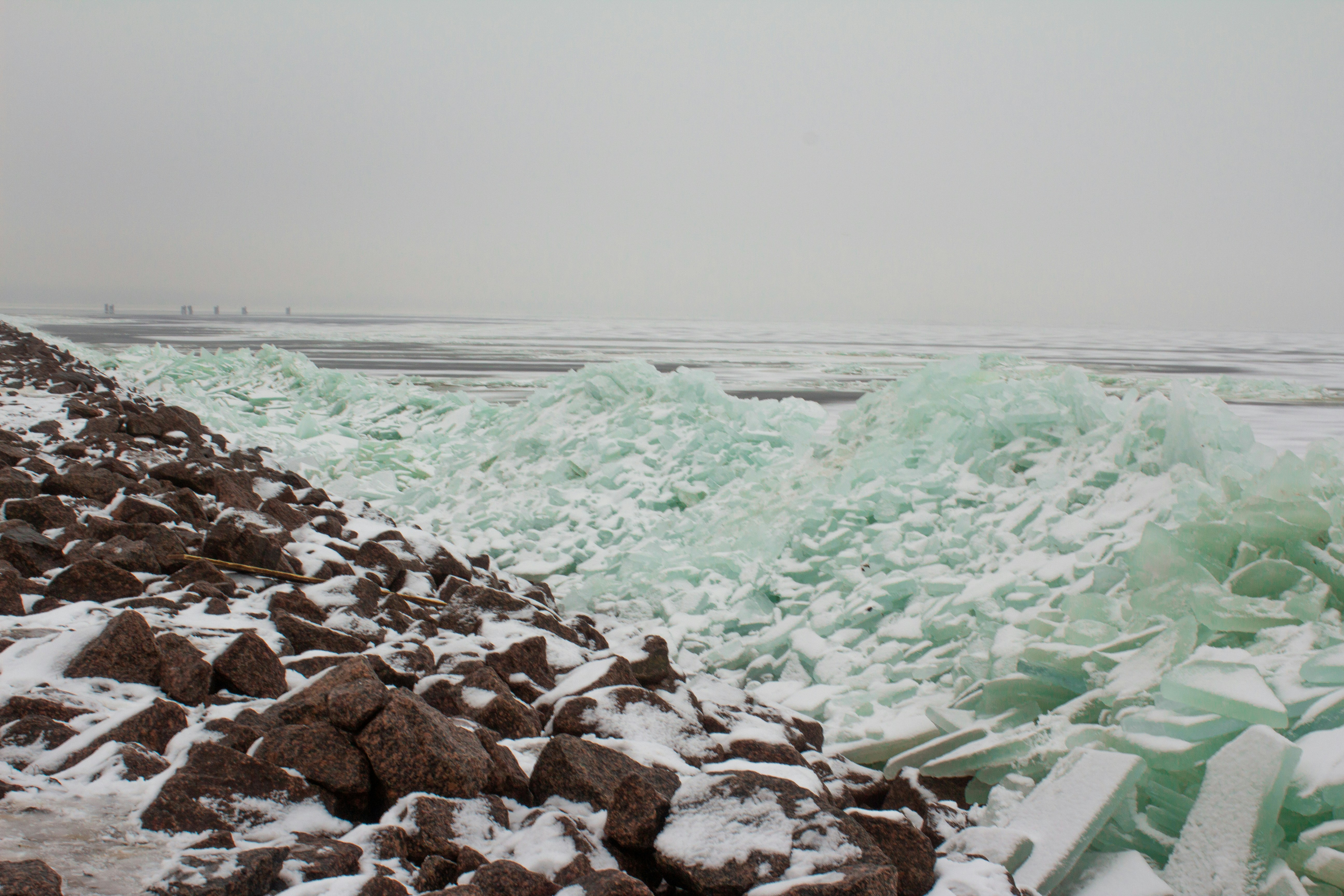 Jagged ice formations rise from a snowy shoreline, contrasting with a muted winter sky. The scene captures the raw beauty of winter's grasp on the landscape.