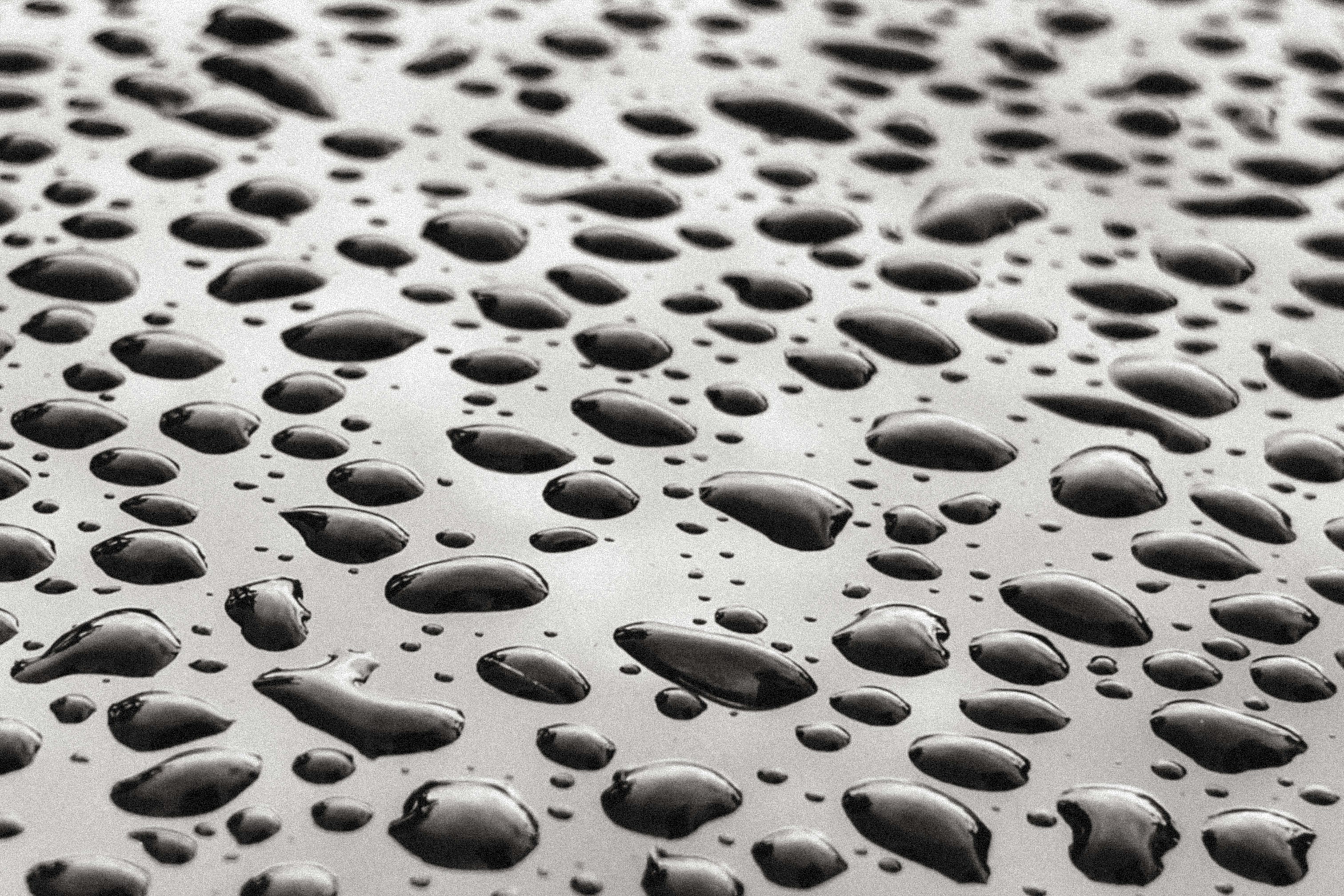 Close-up view of droplets resting on a smooth surface, showcasing their spherical shapes and reflections. The monochrome tones enhance the visual texture.