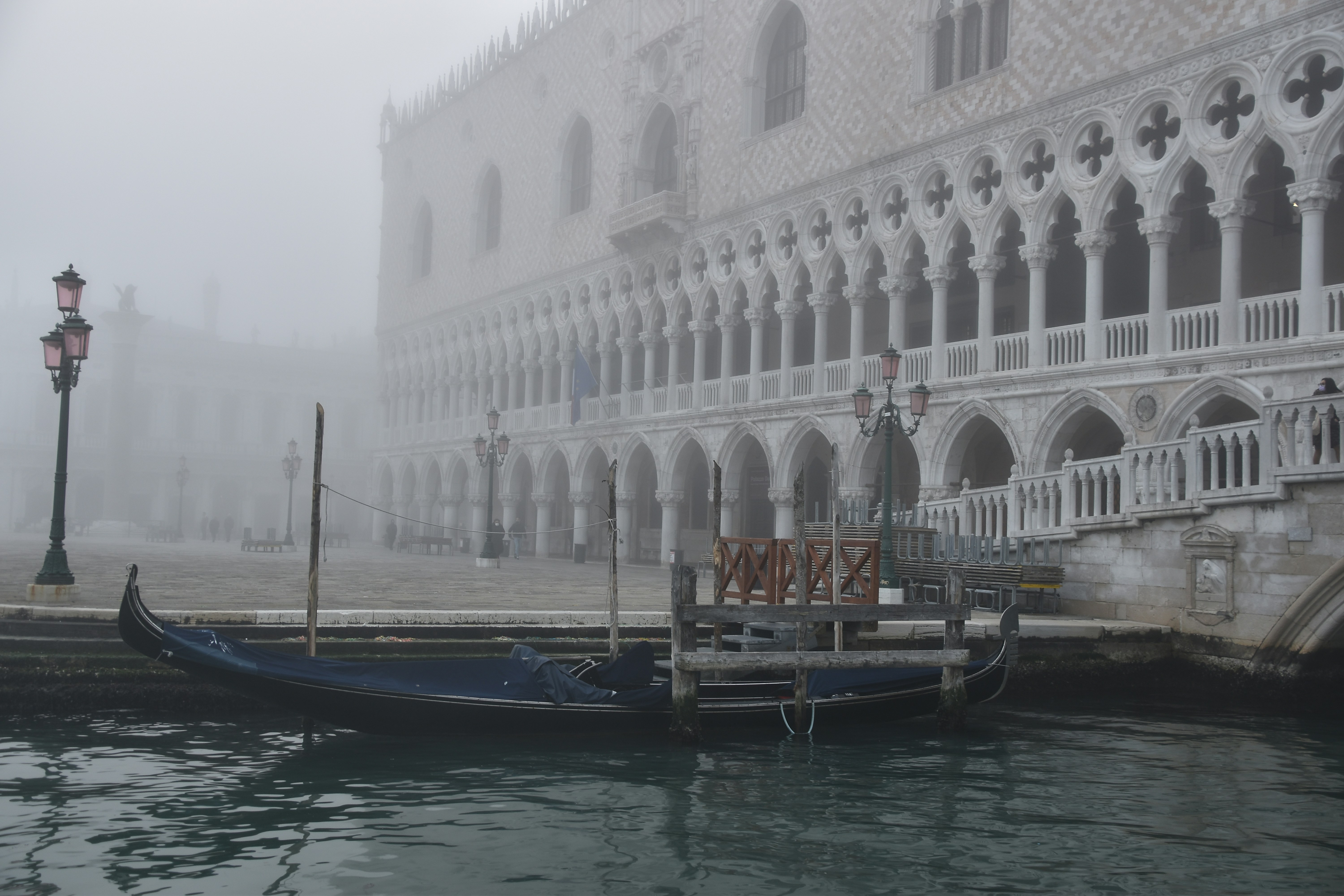 A gondola rests quietly at the edge of a fog-laden canal, framed by the intricate architecture of a historic Venetian building. The scene evokes a sense of serene isolation.