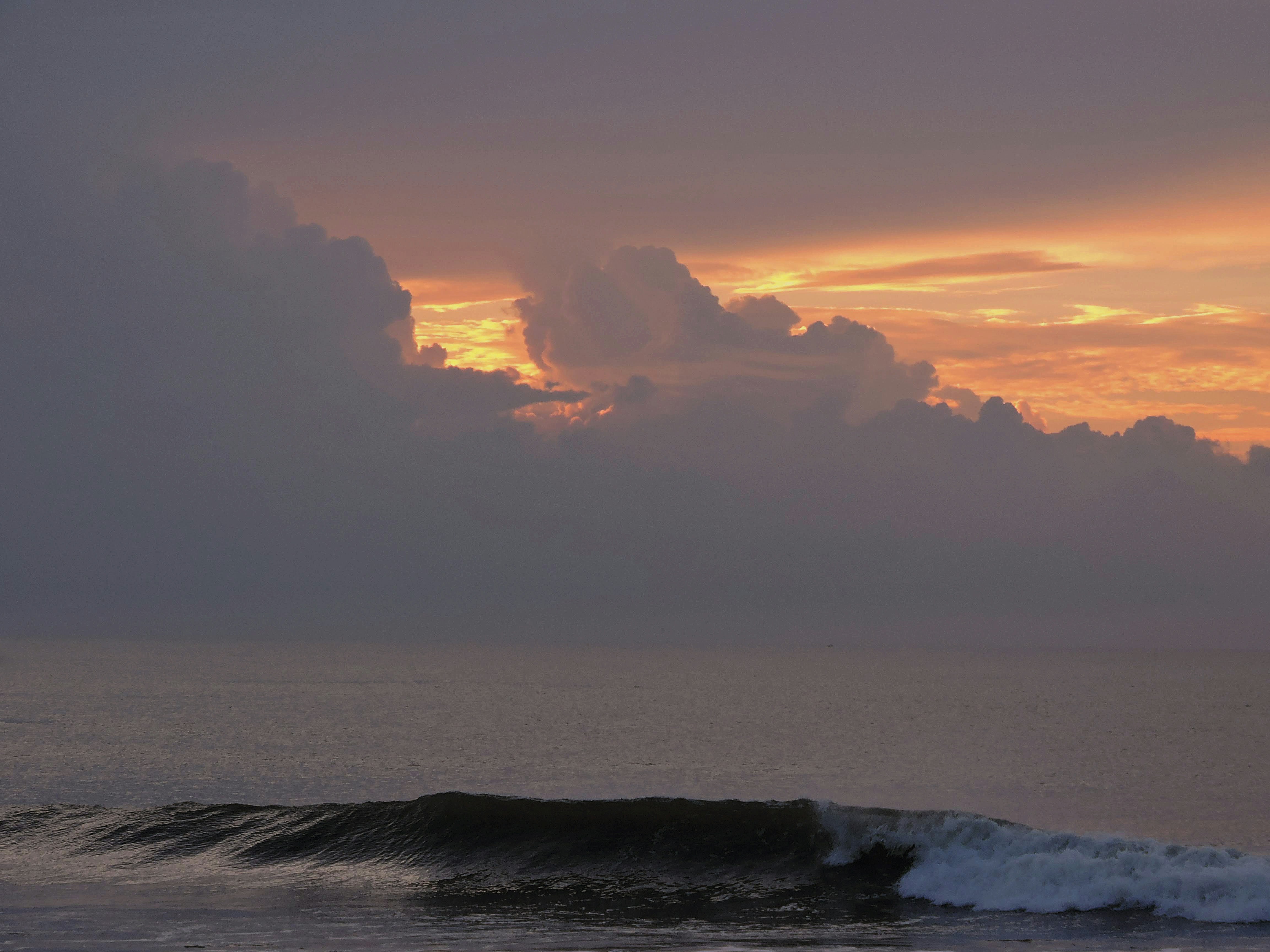 Dusk over a calm sea with a curling foreground wave and a glow of orange light peeking through clouds at the horizon.
