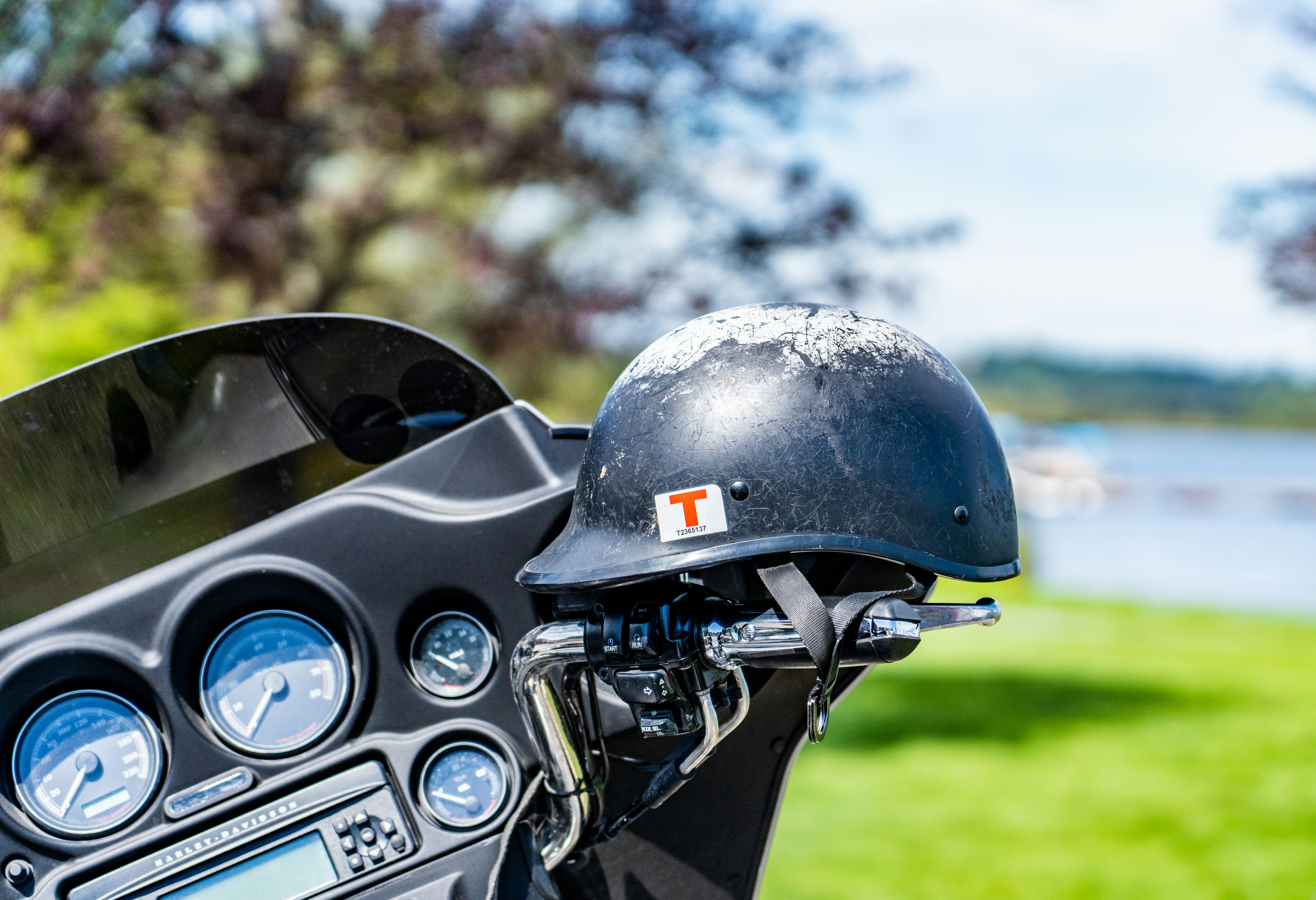 Close-up of a motorcycle's dashboard featuring a weathered helmet with a bold 'T' sticker, set against a vibrant green landscape and distant water.