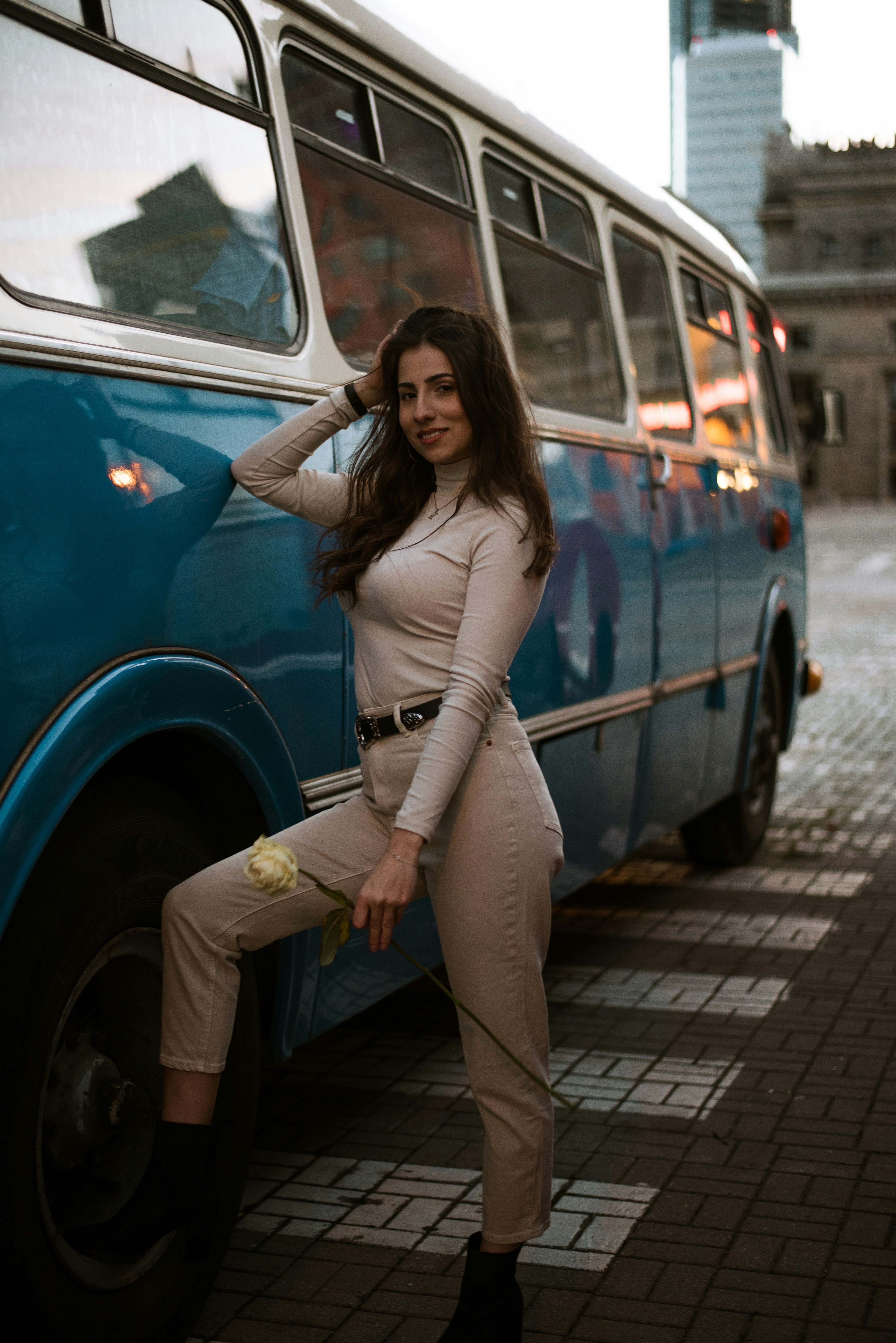 Portrait beside a teal transit giant: a poised figure with a white turtleneck and flower