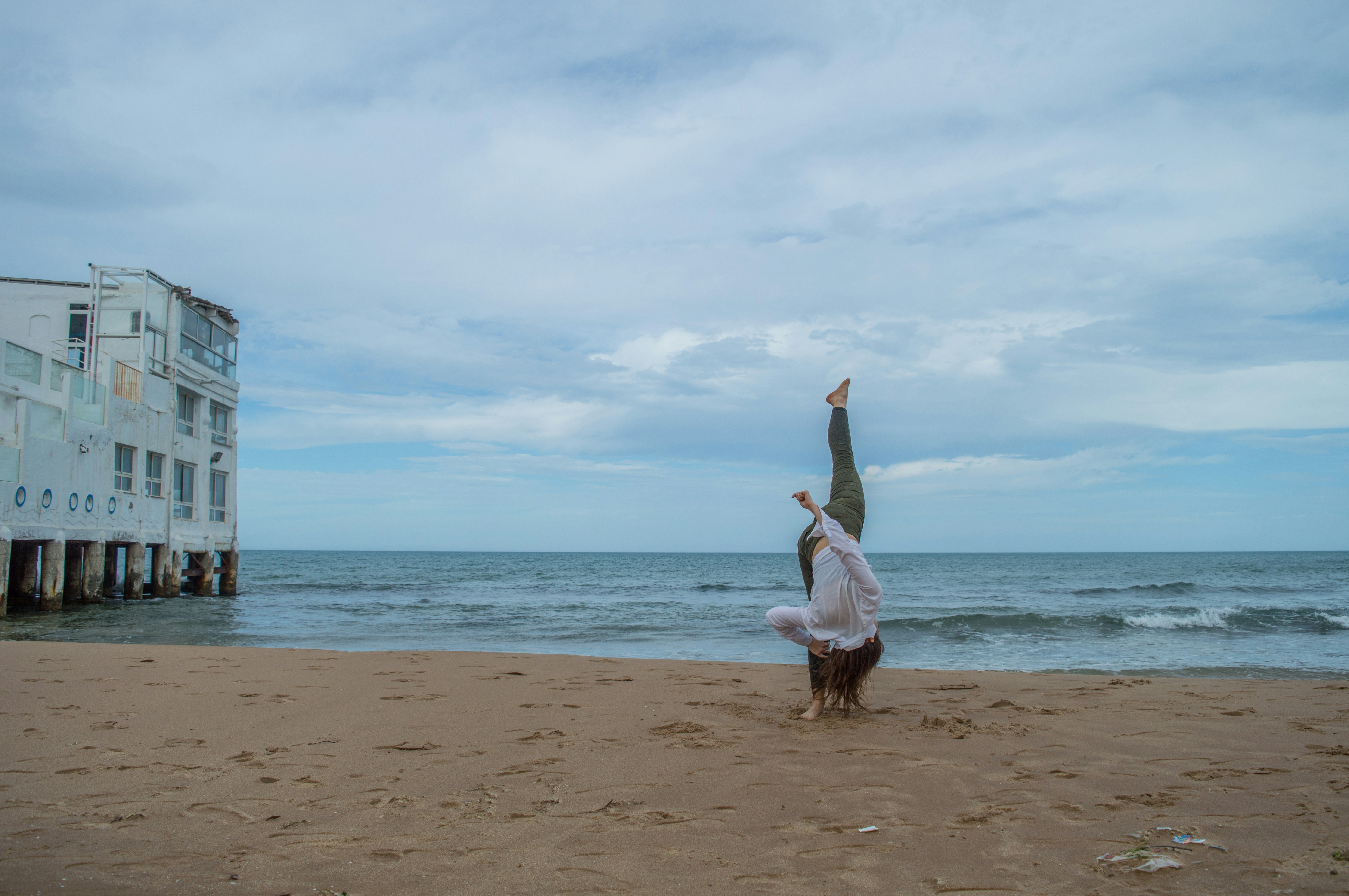 Person performing a handstand on a sandy beach with a building on stilts by the sea under a cloudy sky.