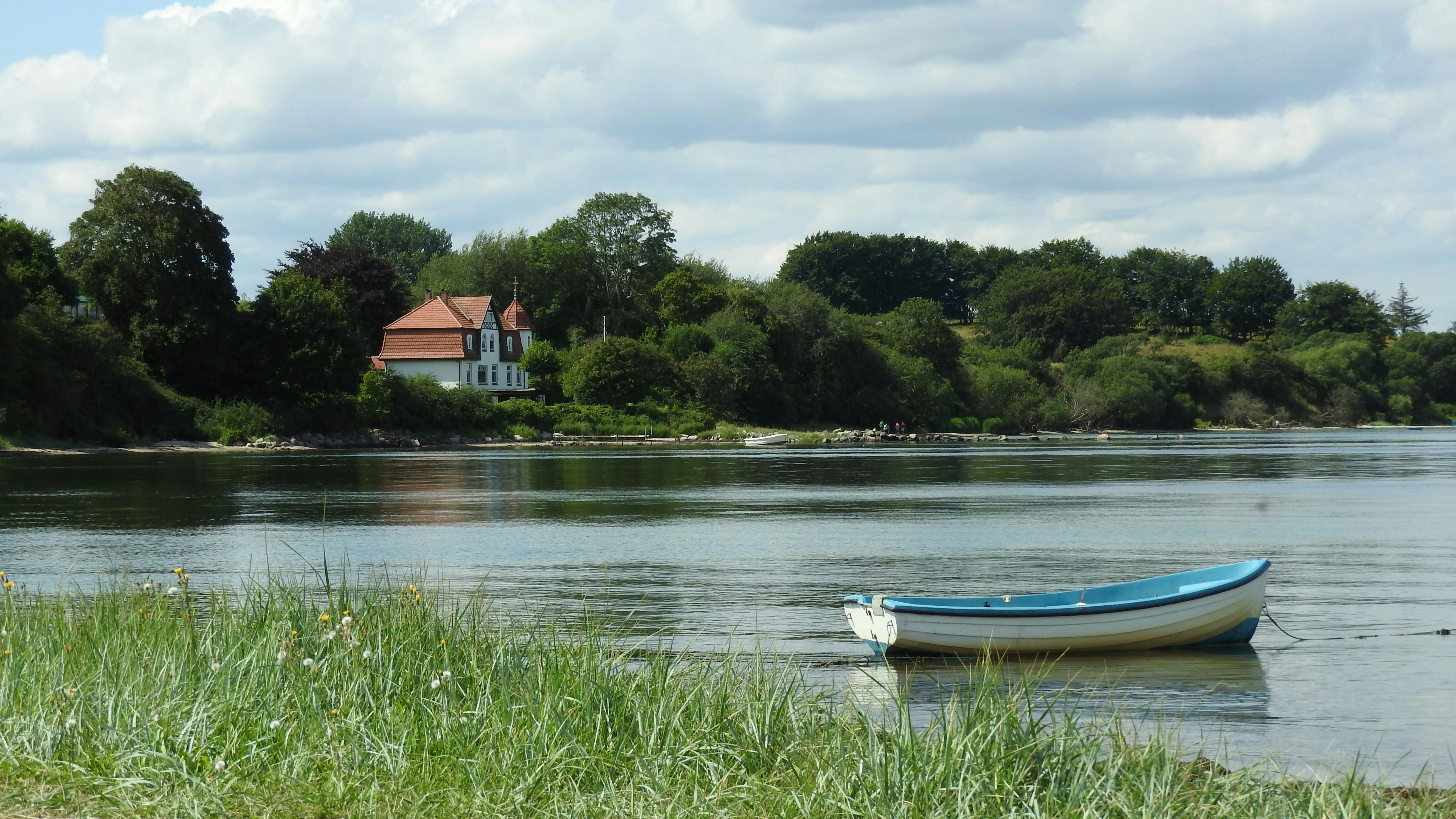 Small boat floats peacefully on a calm lake with lush greenery and a cottage in the background.