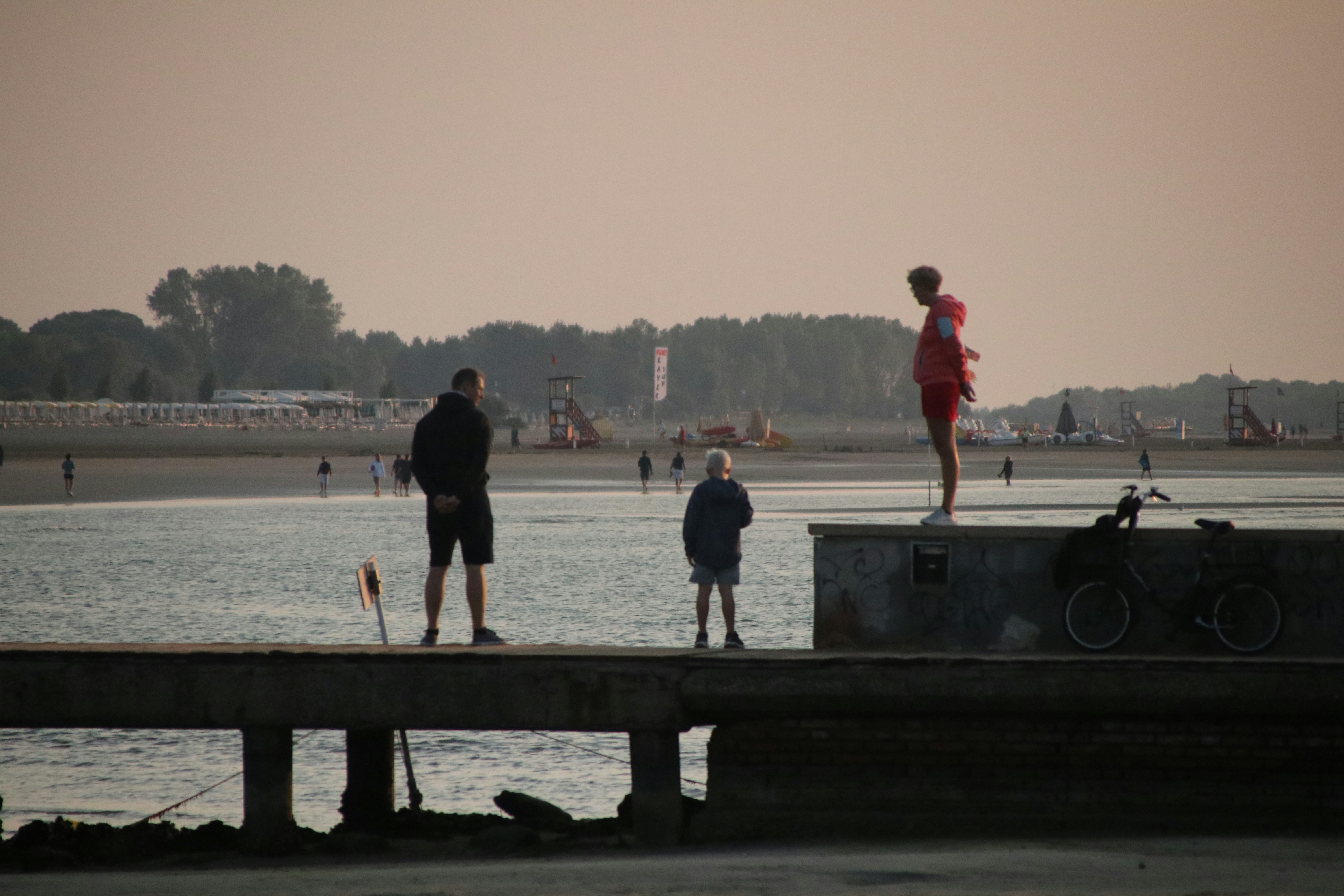 Three figures stand near the water's edge, with one person perched on a ledge, creating a serene atmosphere at dusk.