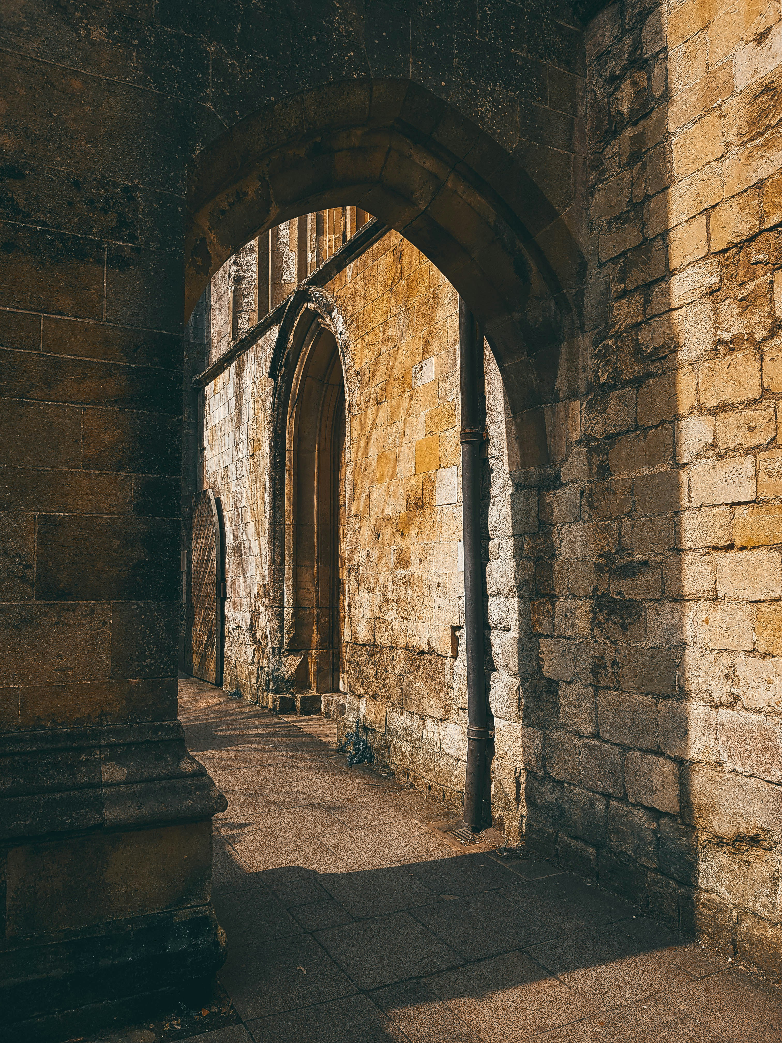 Ancient stone archway framing a narrow pathway leading to a weathered entrance, illuminated by soft sunlight. 