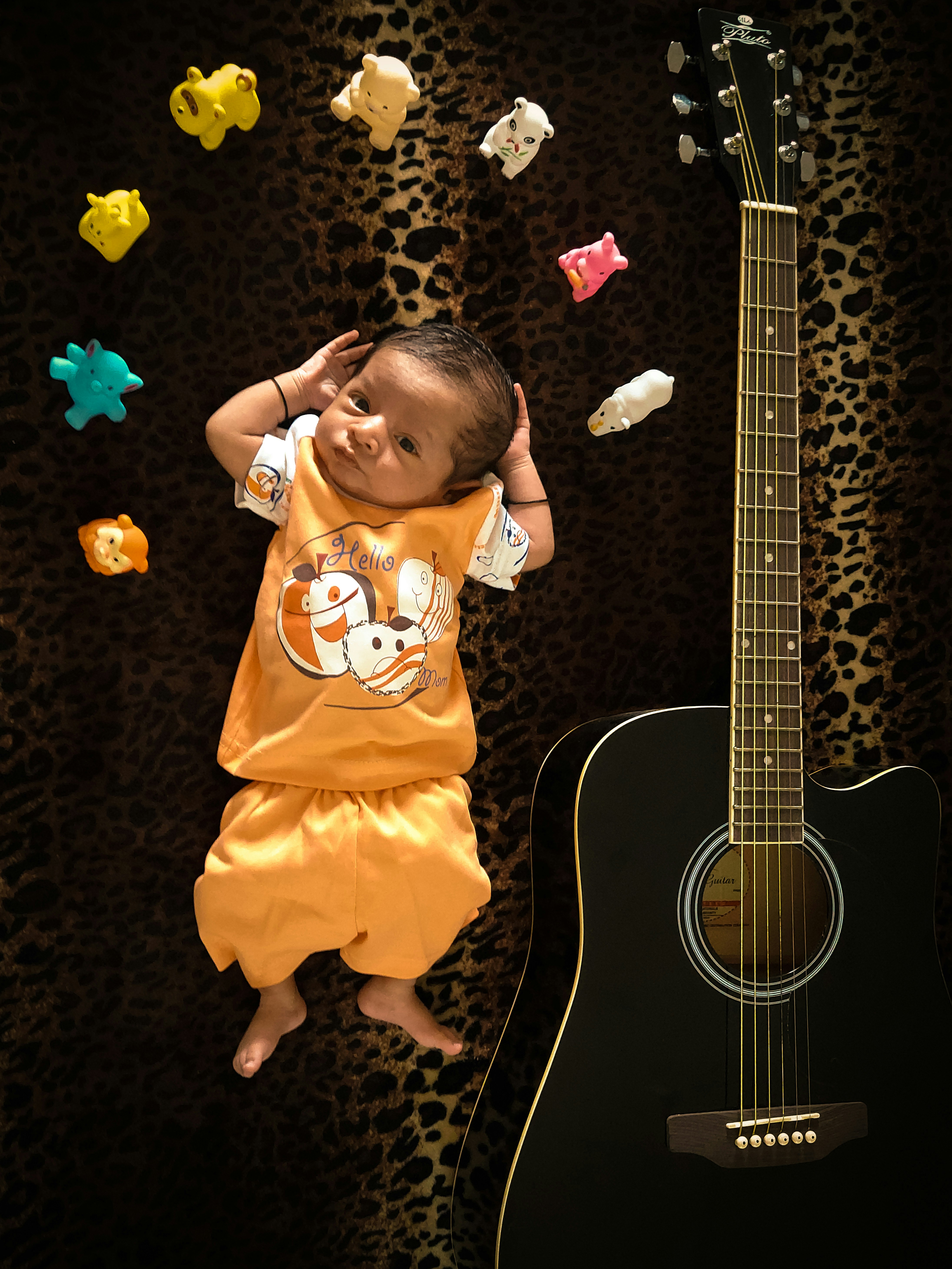 A baby in an orange outfit rests on a leopard print background, surrounded by colorful toys and a guitar. The scene captures a whimsical blend of childhood and music.
