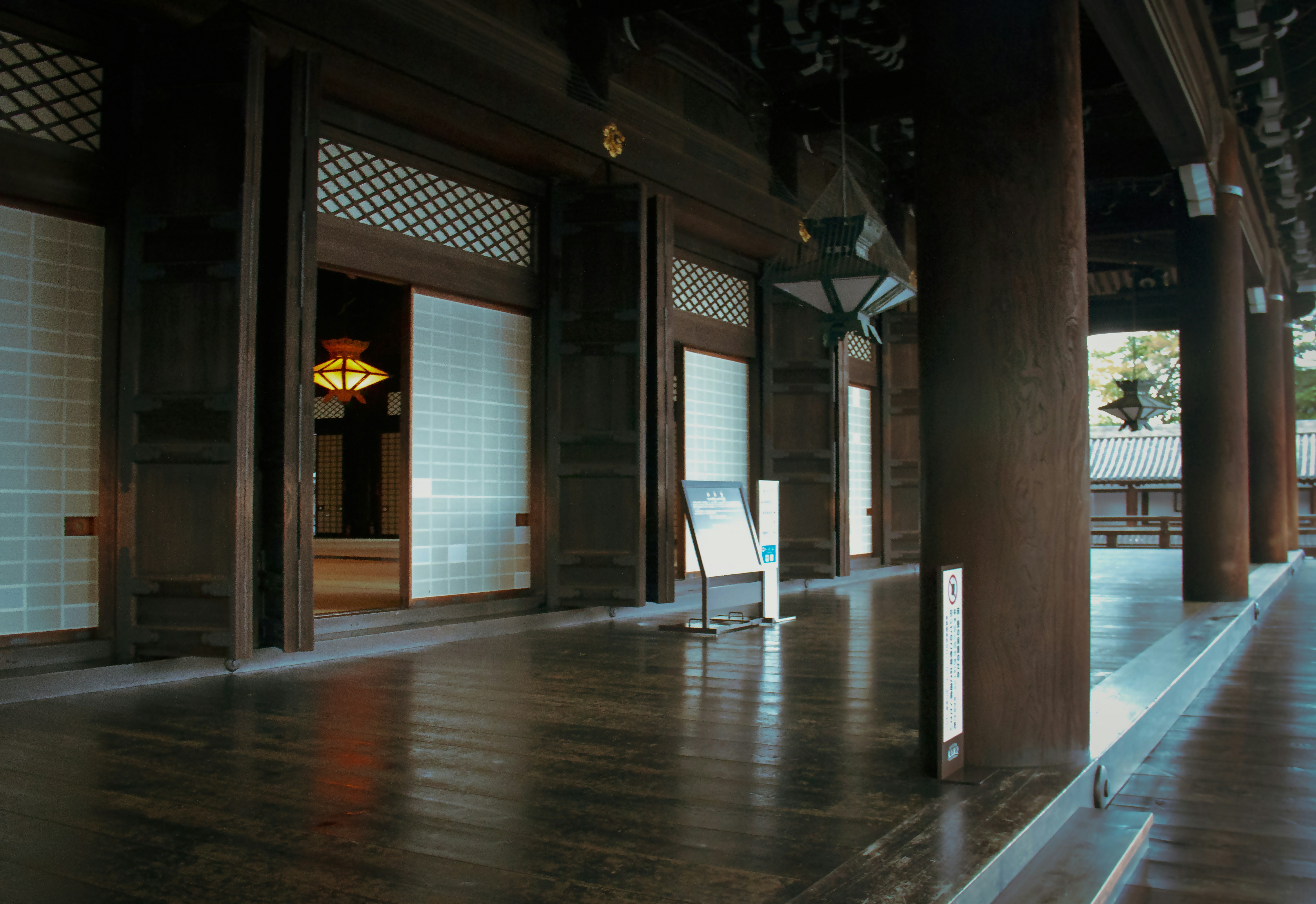Interior of a traditional building showcasing wooden pillars and sliding doors, illuminated by soft light from lanterns. A sign stands in the foreground.