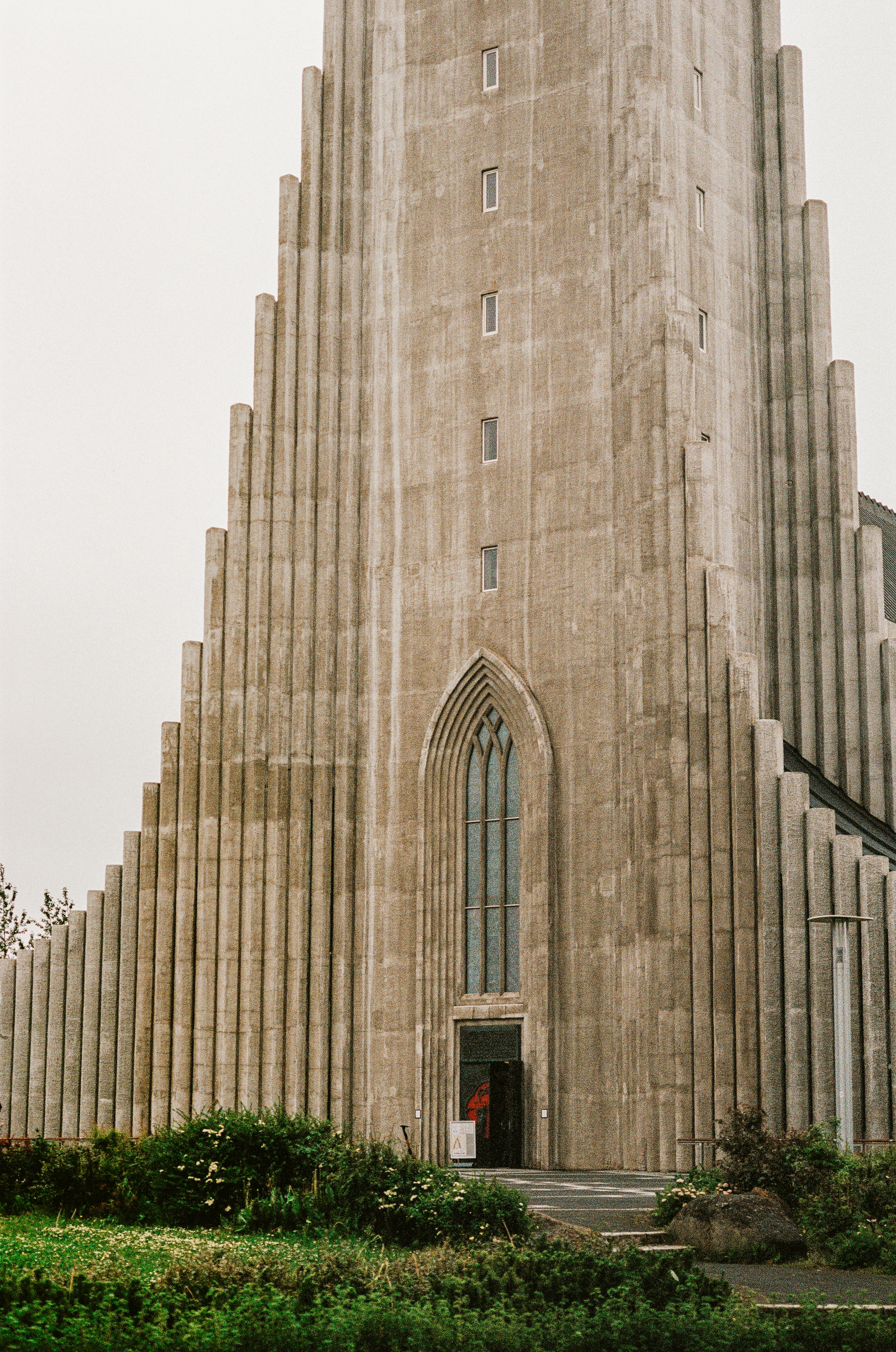 Concrete structure with sharp angular lines and a prominent entrance, showcasing modernist design principles. Lush greenery at the base adds a touch of nature.