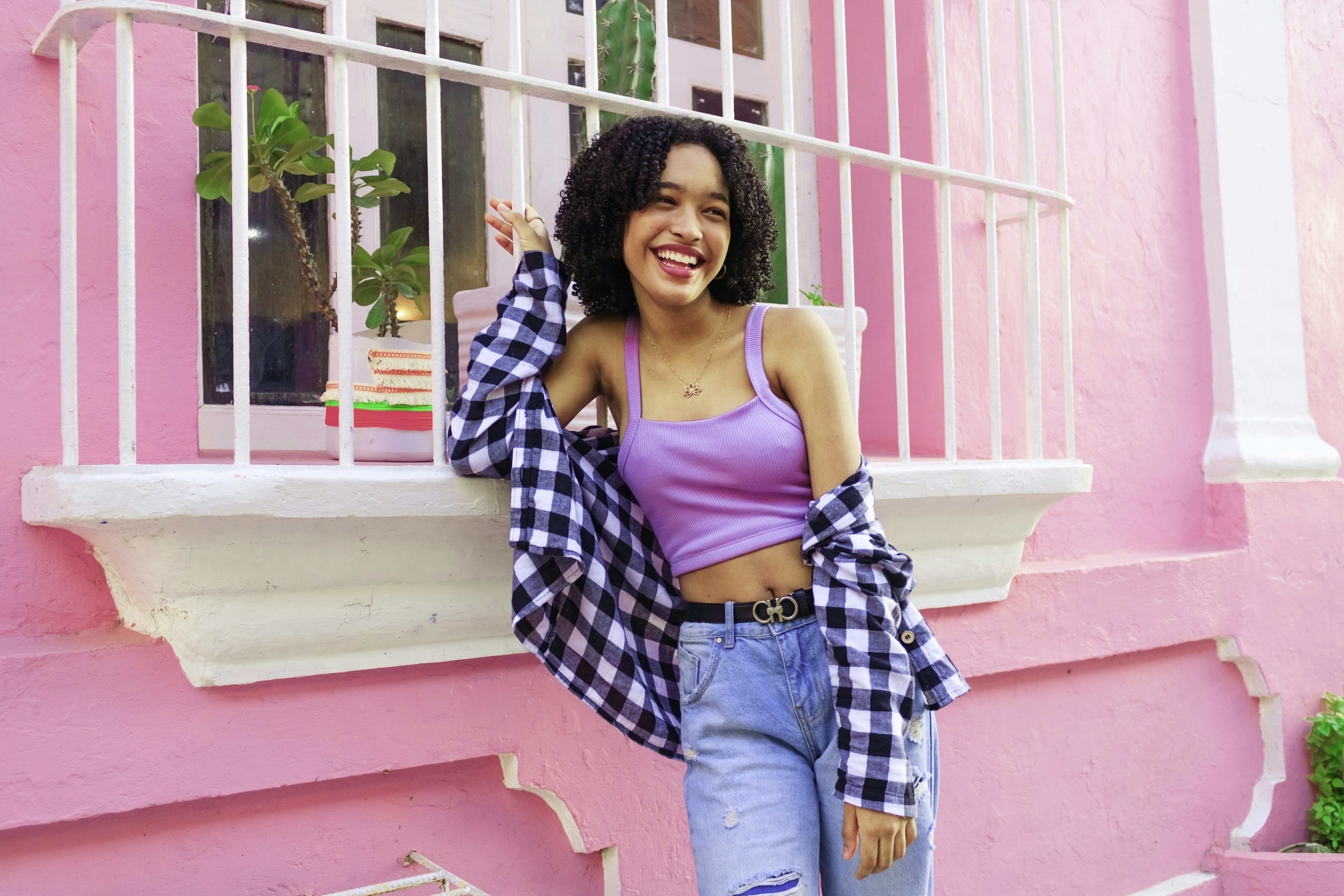 A young woman with curly hair smiles while leaning against a pink wall, wearing a purple top and a checkered shirt. The scene captures a lively urban atmosphere.