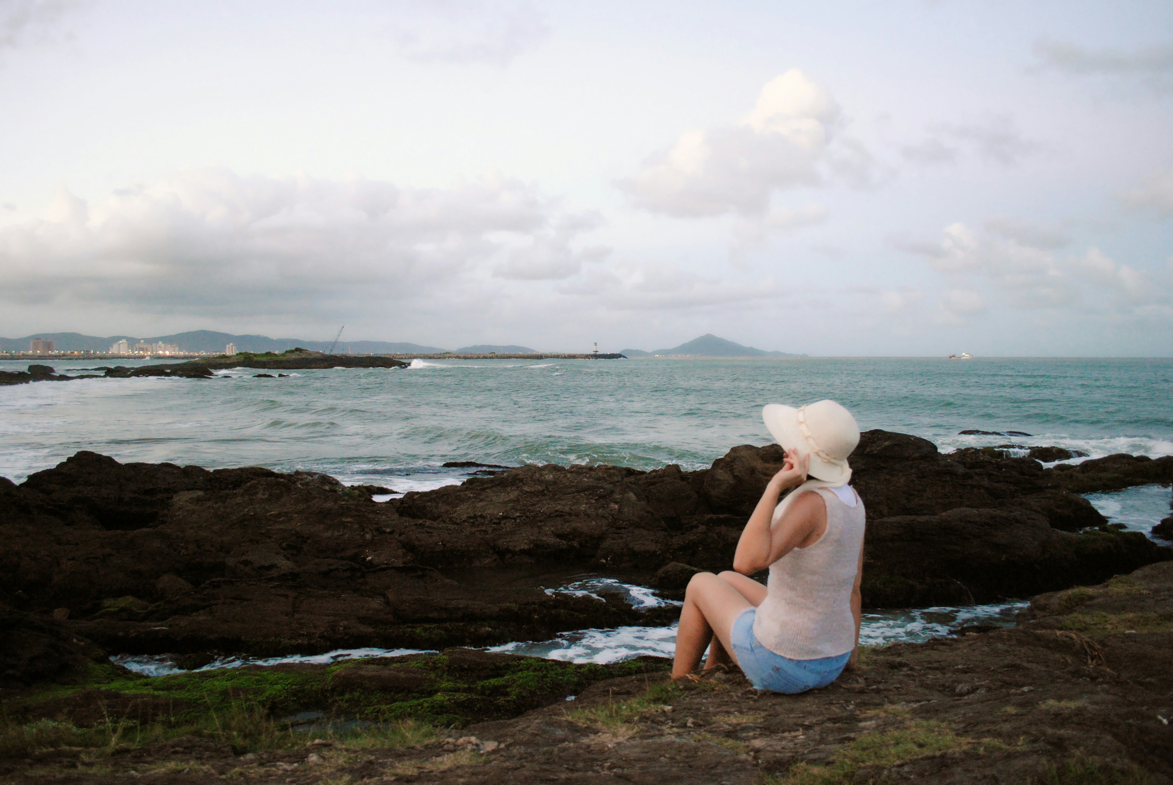 A woman in a wide-brimmed hat sits on rocky shores, gazing thoughtfully at the ocean waves under a soft sky. The scene captures a moment of tranquility and reflection.