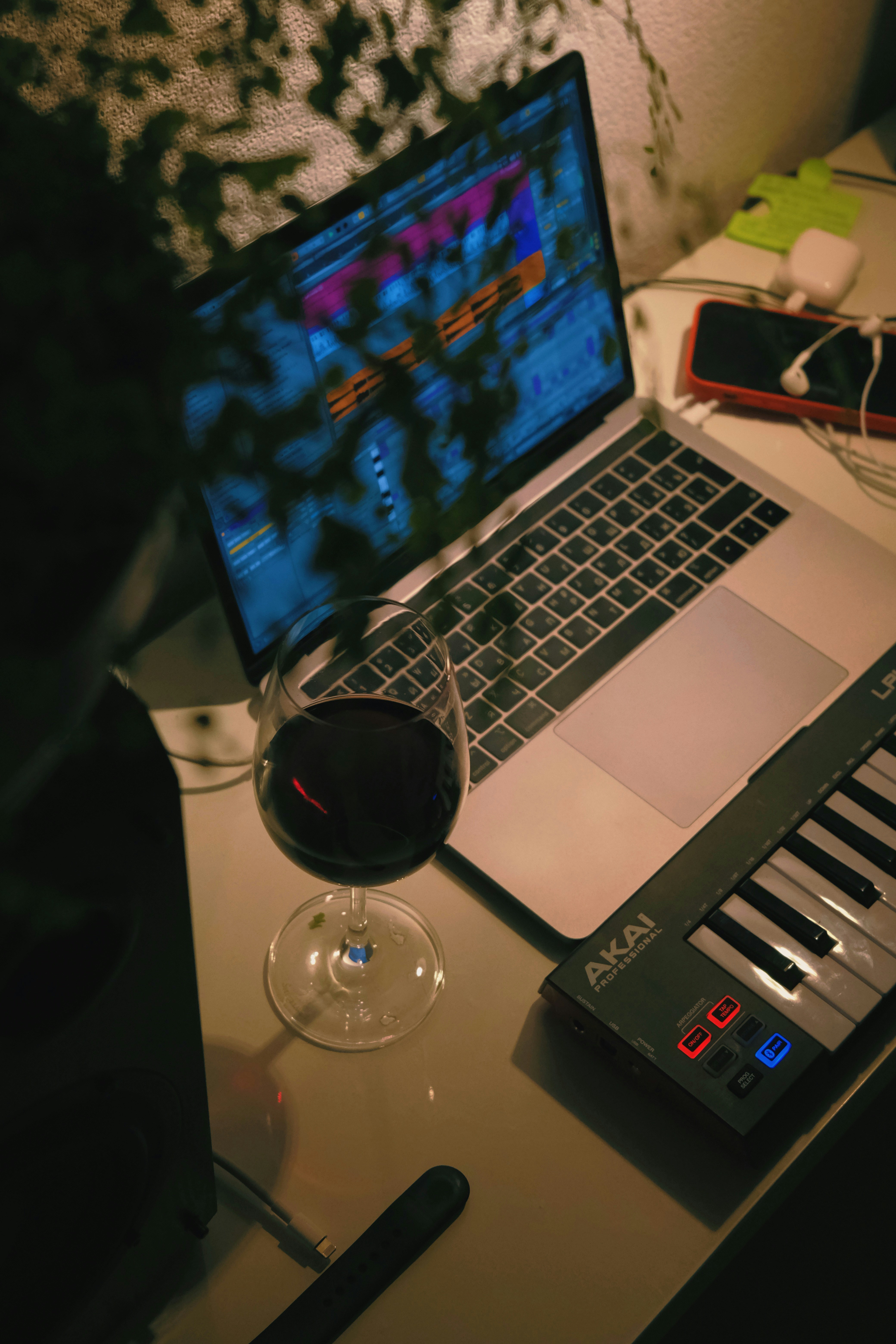 A laptop displaying music software sits on a desk alongside an AKAI keyboard and a glass of red wine, creating an inviting atmosphere for creativity.