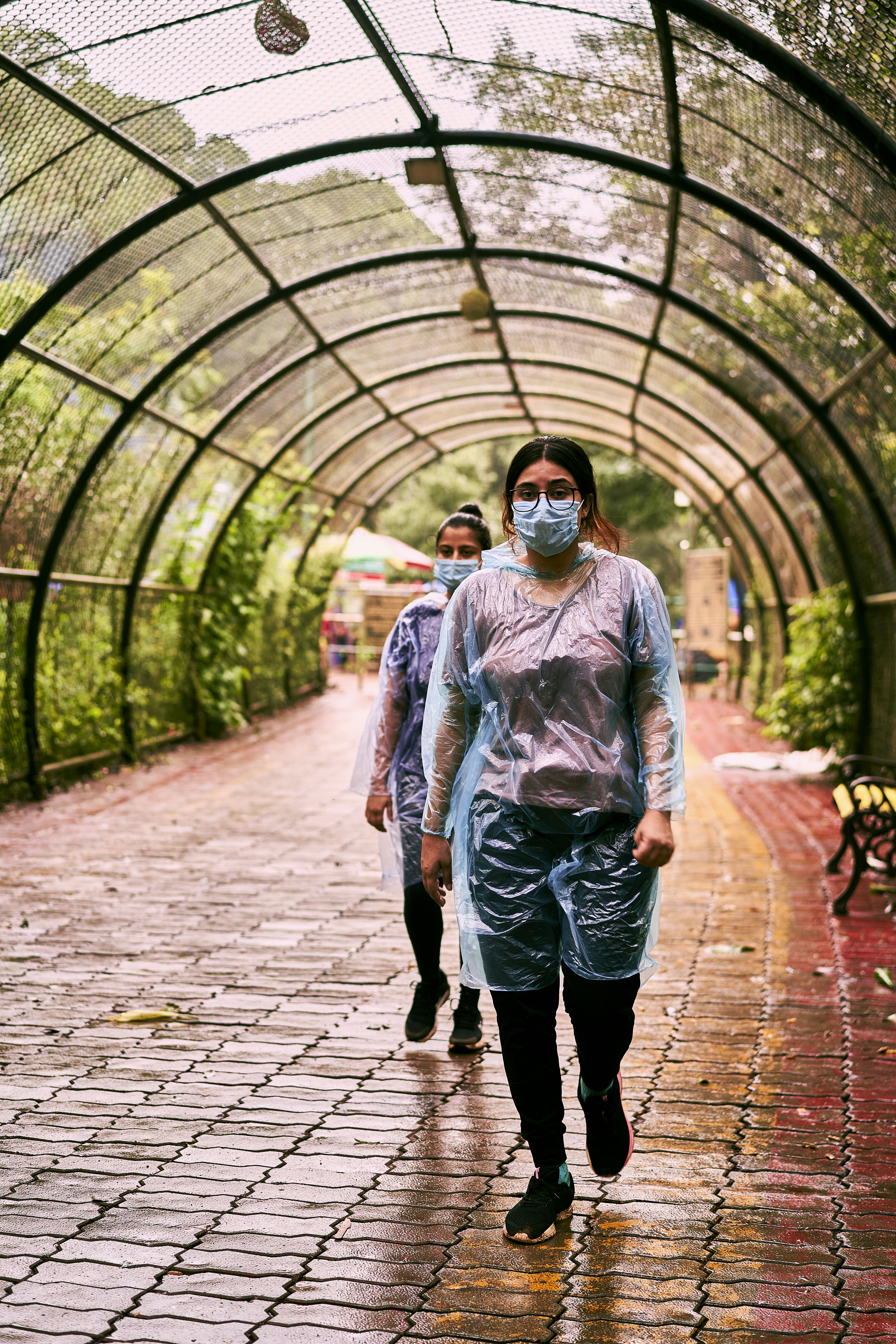 Two individuals in rain gear walk along a wet pathway under a canopy of greenery. The scene captures the essence of perseverance in a rainy environment.