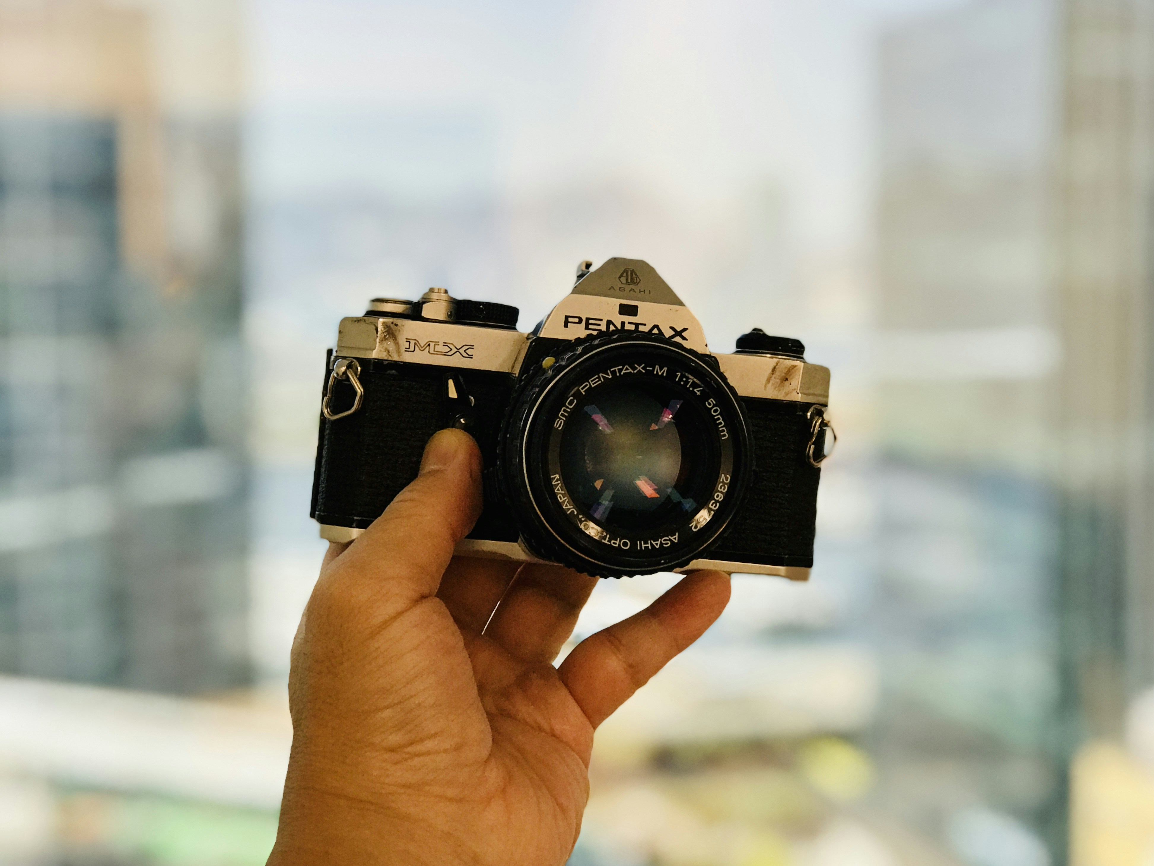 A hand holds a vintage Pentax MX camera against a blurred urban backdrop, highlighting its classic design and lens details.