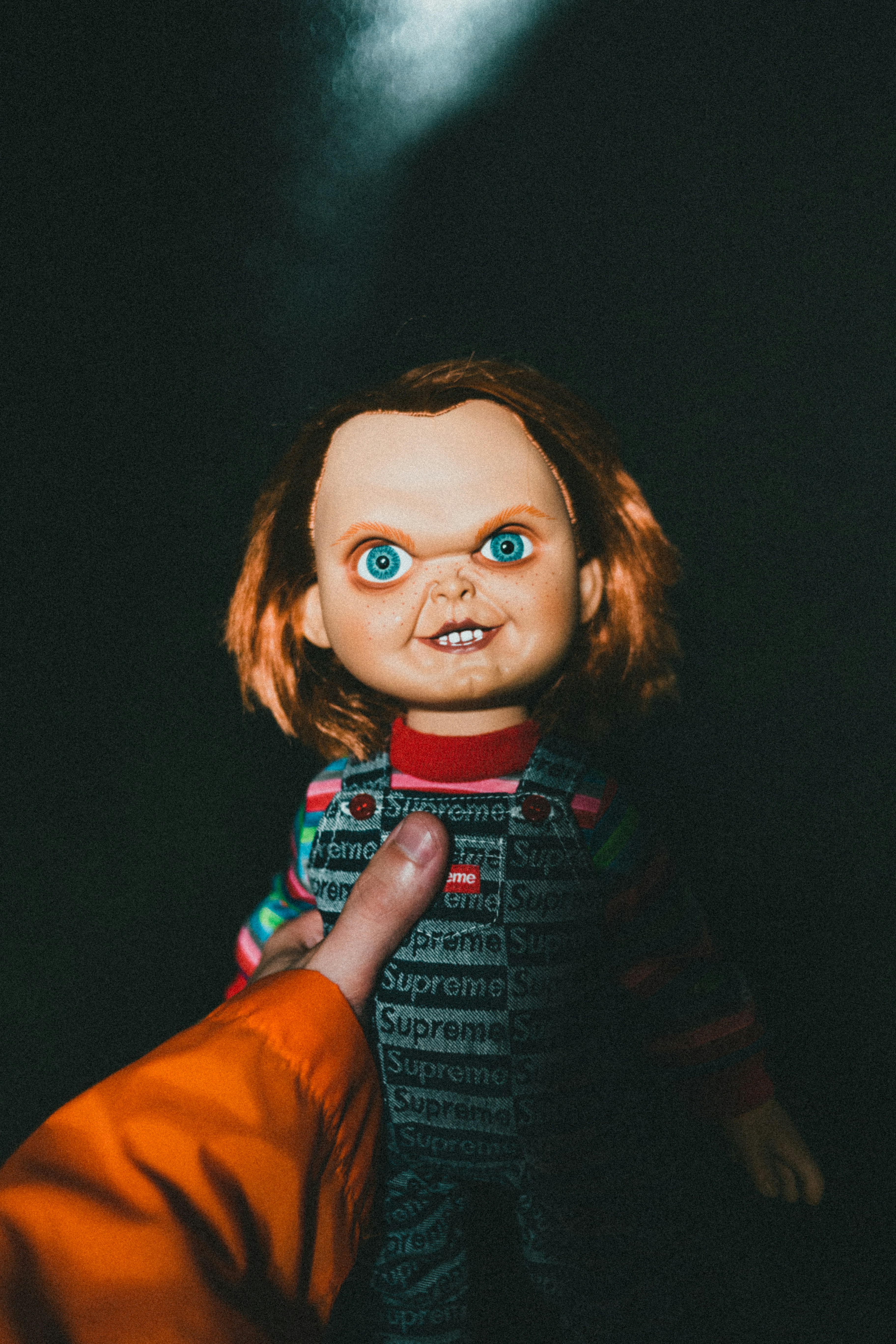 A close-up of a Chucky doll with striking blue eyes, held by a hand in a brightly colored jacket against a dark background.